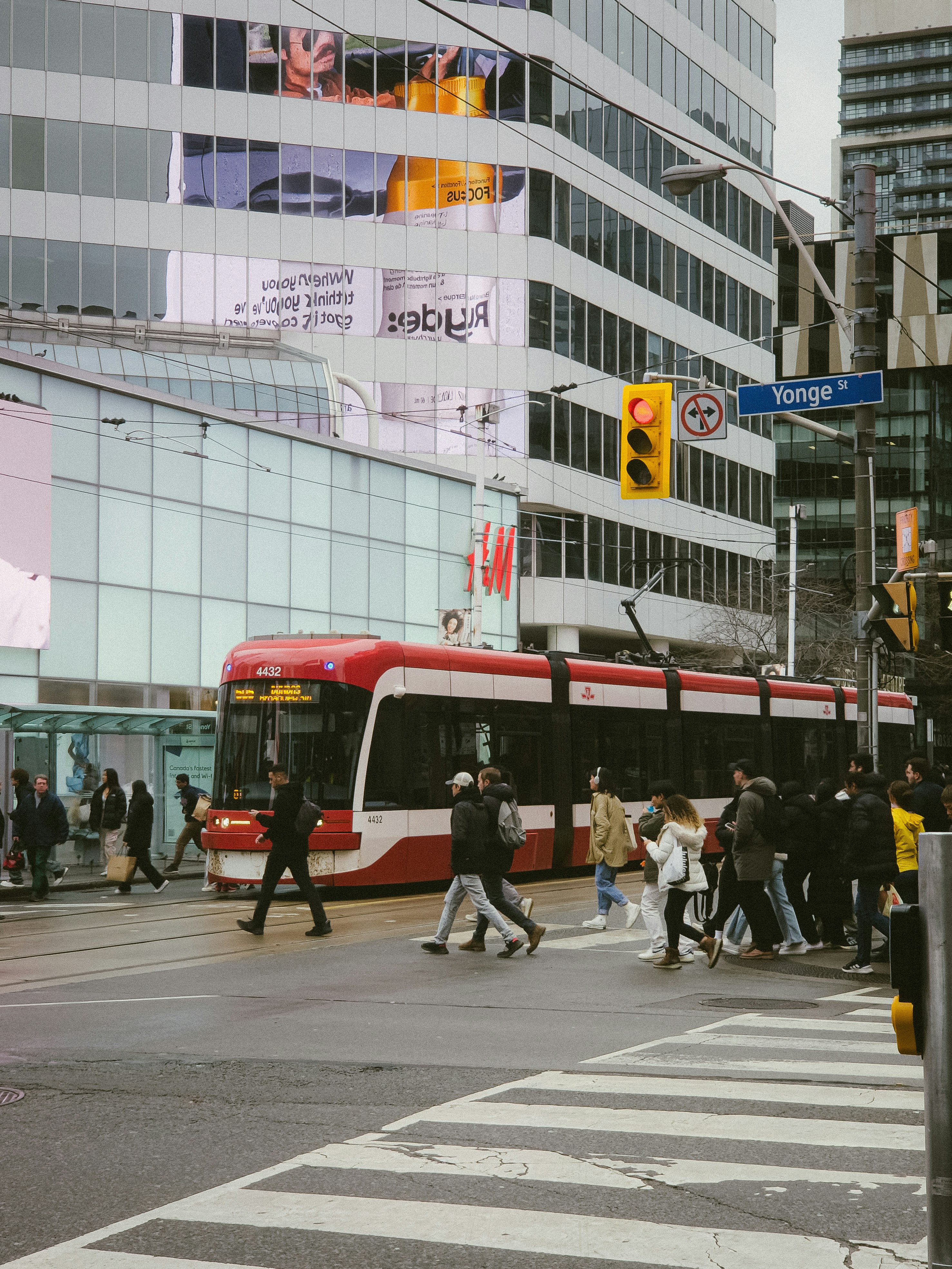 A busy city street scene featuring a red-and-white tram crossing a zebra-striped crosswalk as pedestrians pass beneath glass-fronted towers.