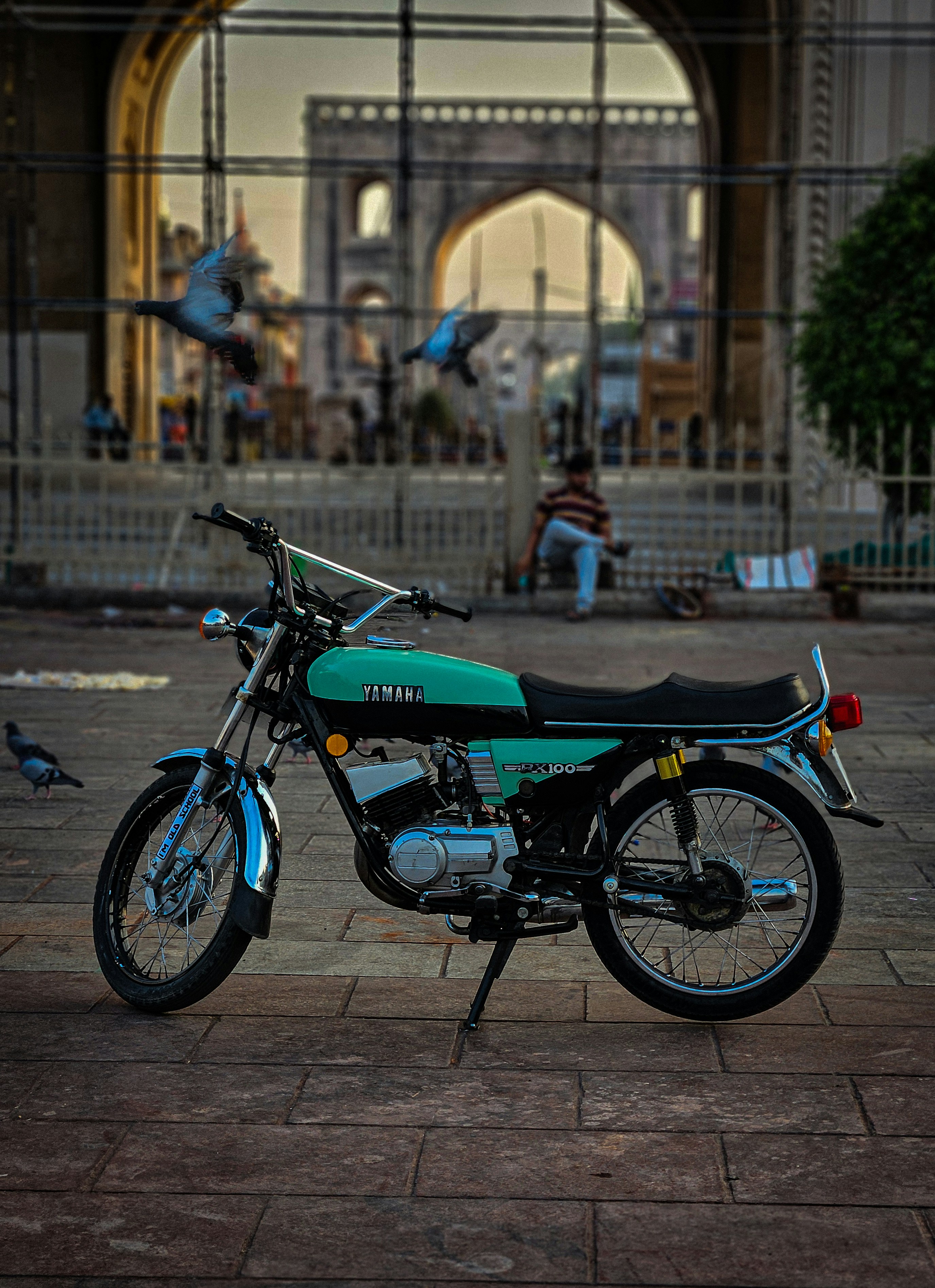 Classic Yamaha motorcycle parked in front of an architectural landmark, with pigeons in motion and people in the background.