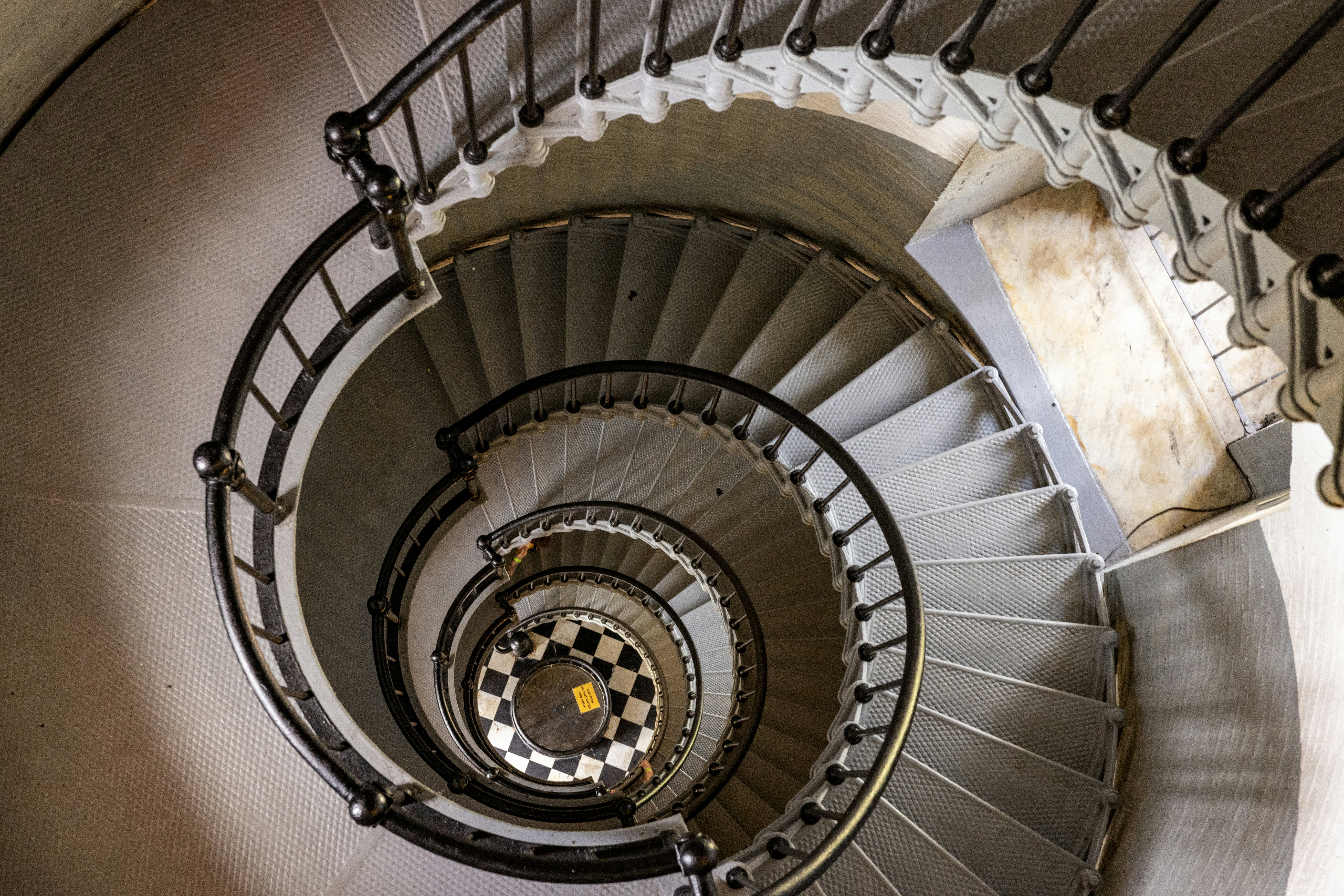 Spiral staircase inside a lighthouse, viewed from above, with intricate railing details and a checkerboard floor at the base.