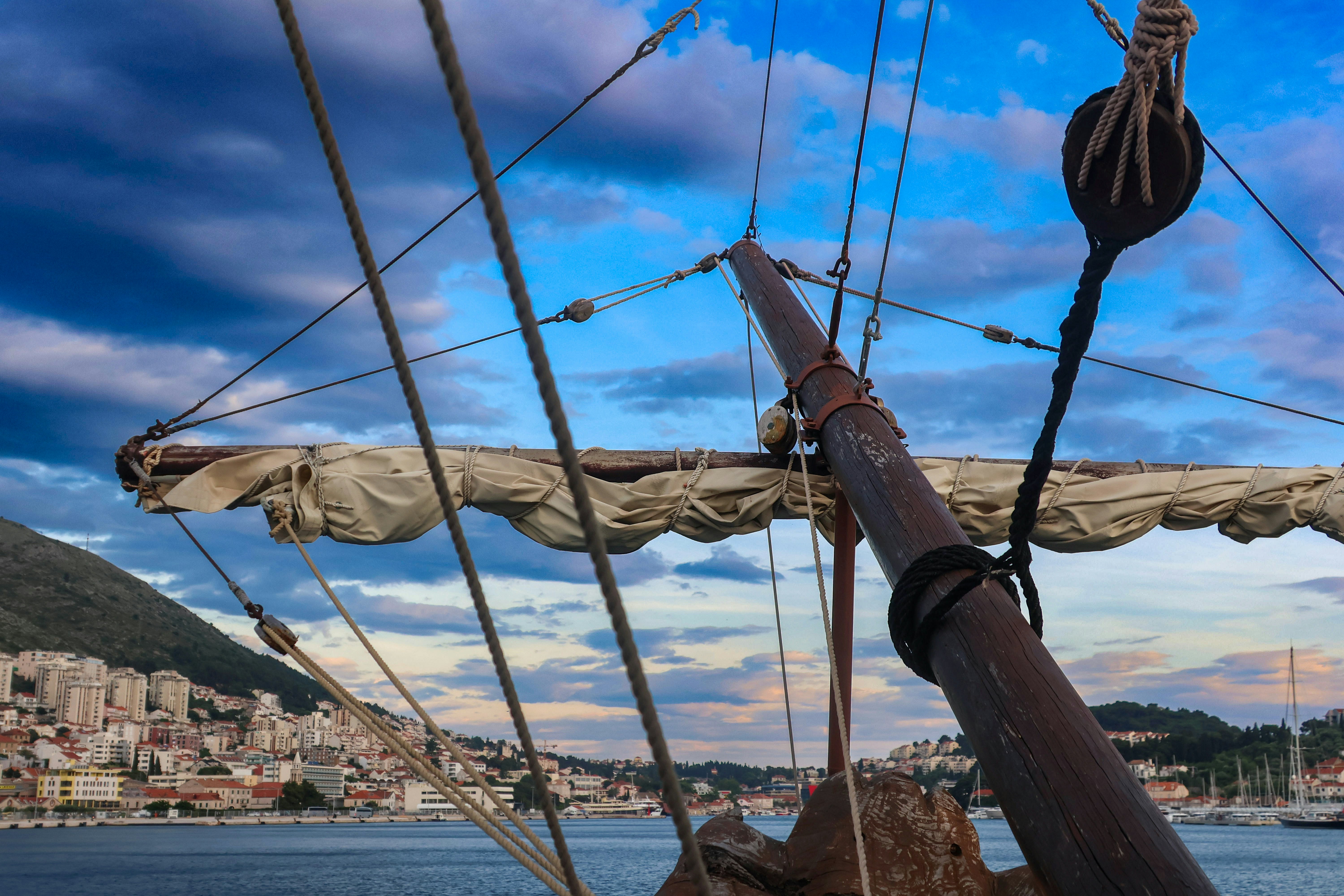 This captivating image captures the intricate rigging of a sailboat set against a vibrant blue sky, with a picturesque coastal town in the background. The interplay of the soft, warm hues of the town and the deep blue sky creates a striking contrast, while the diagonal lines of the ropes and mast add dynamic tension to the composition. The scene evokes a sense of adventure and tranquility, enhanced by the natural lighting and expansive sky.