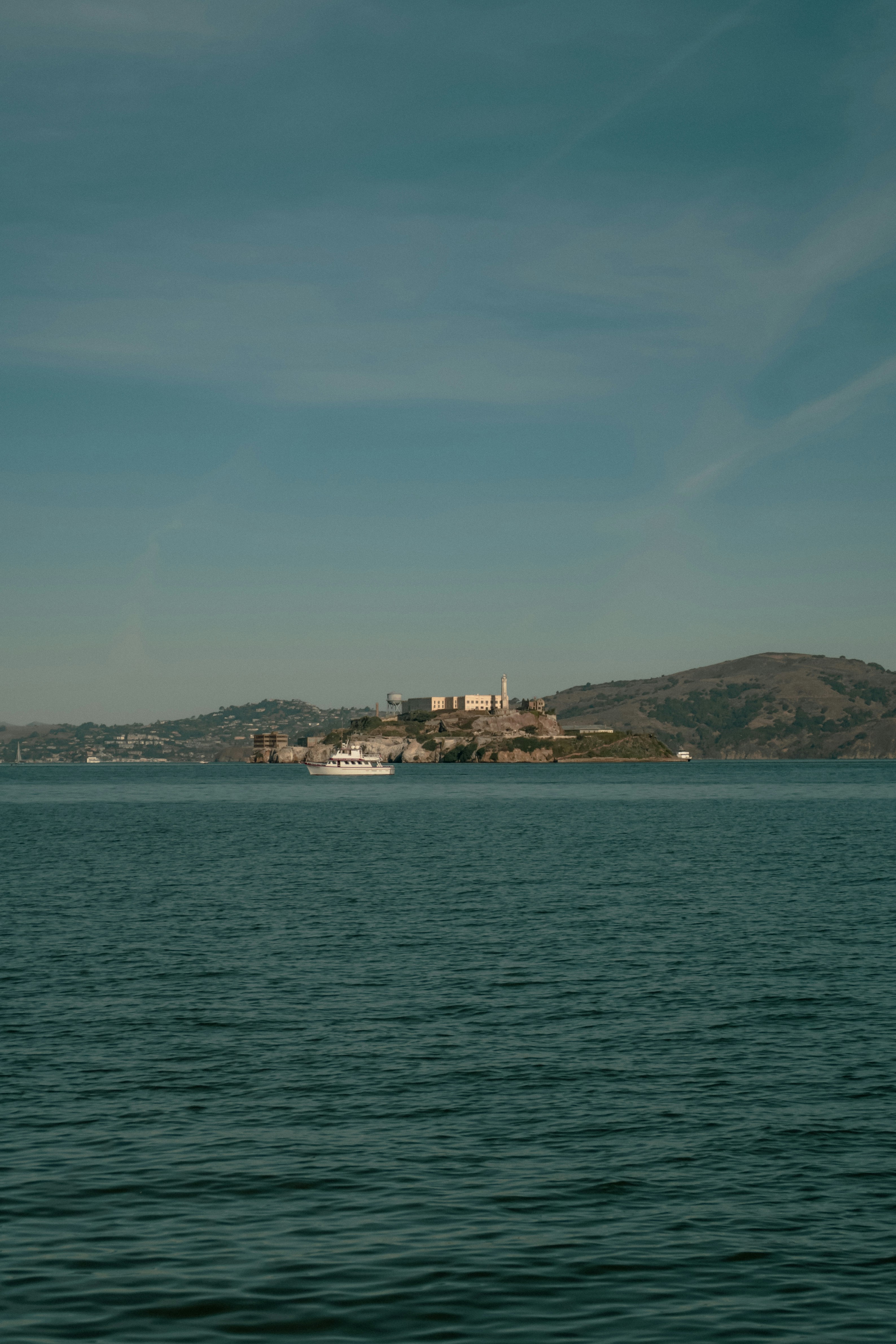 Alcatraz Island stands prominently against a serene bay, with a distant ferry navigating the tranquil waters. The landscape captures a moment of calm amidst the historical significance of the site.