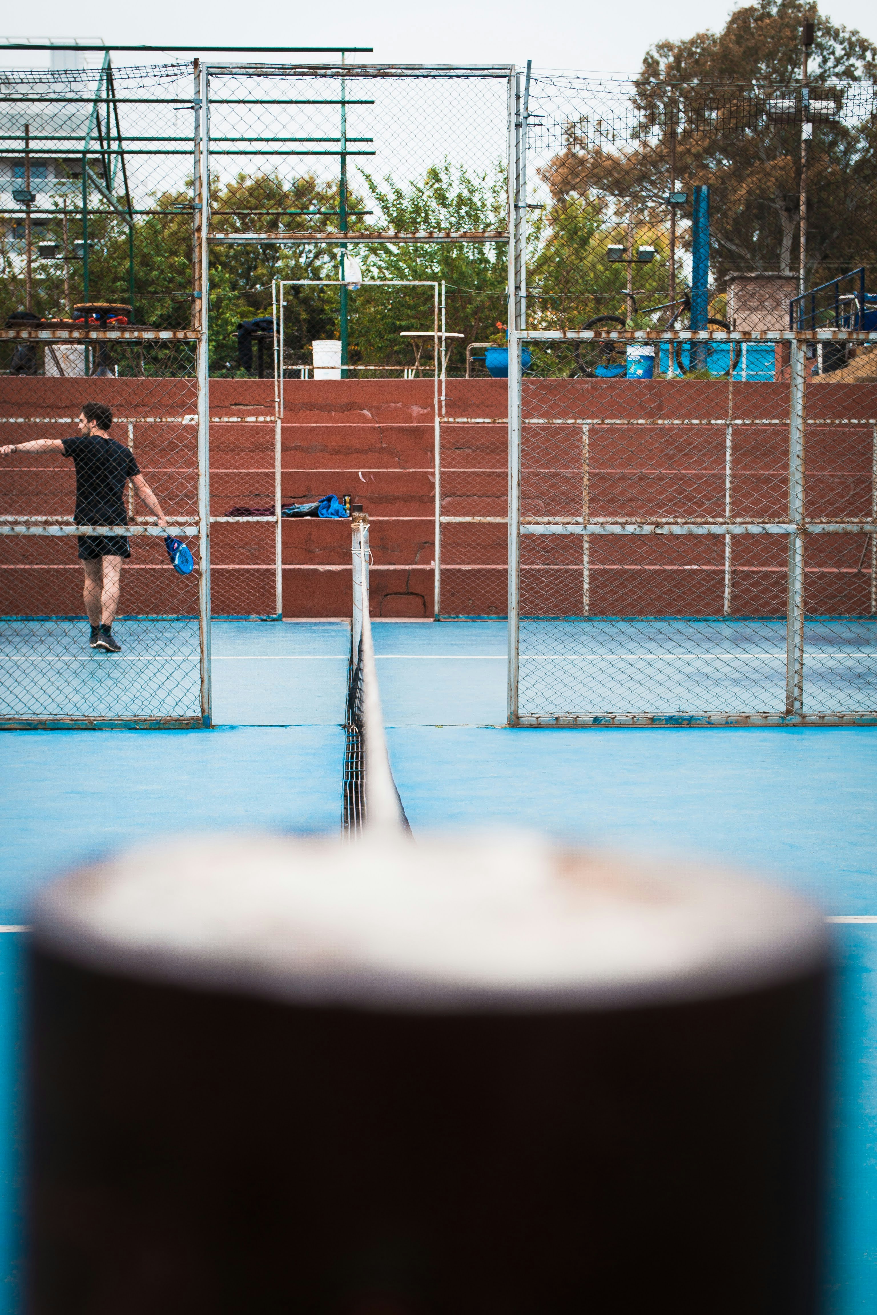 Hands on the Court: A lone player at the net framed by chain-link fences