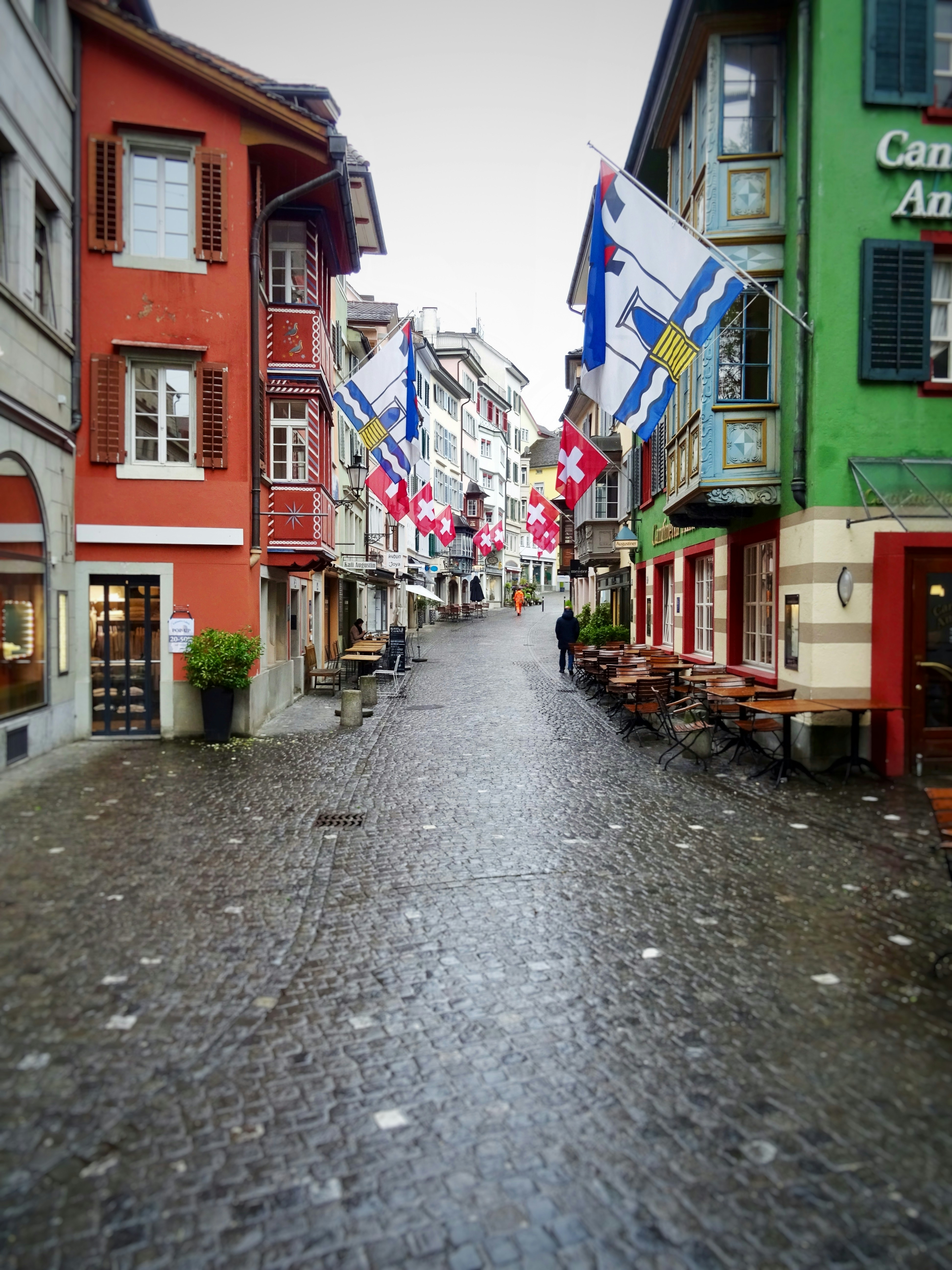 Photograph of a colorful cobblestone street flanked by vibrantly painted buildings and hanging flags. A distant figure at the center enhances the sense of depth toward the town's far end.