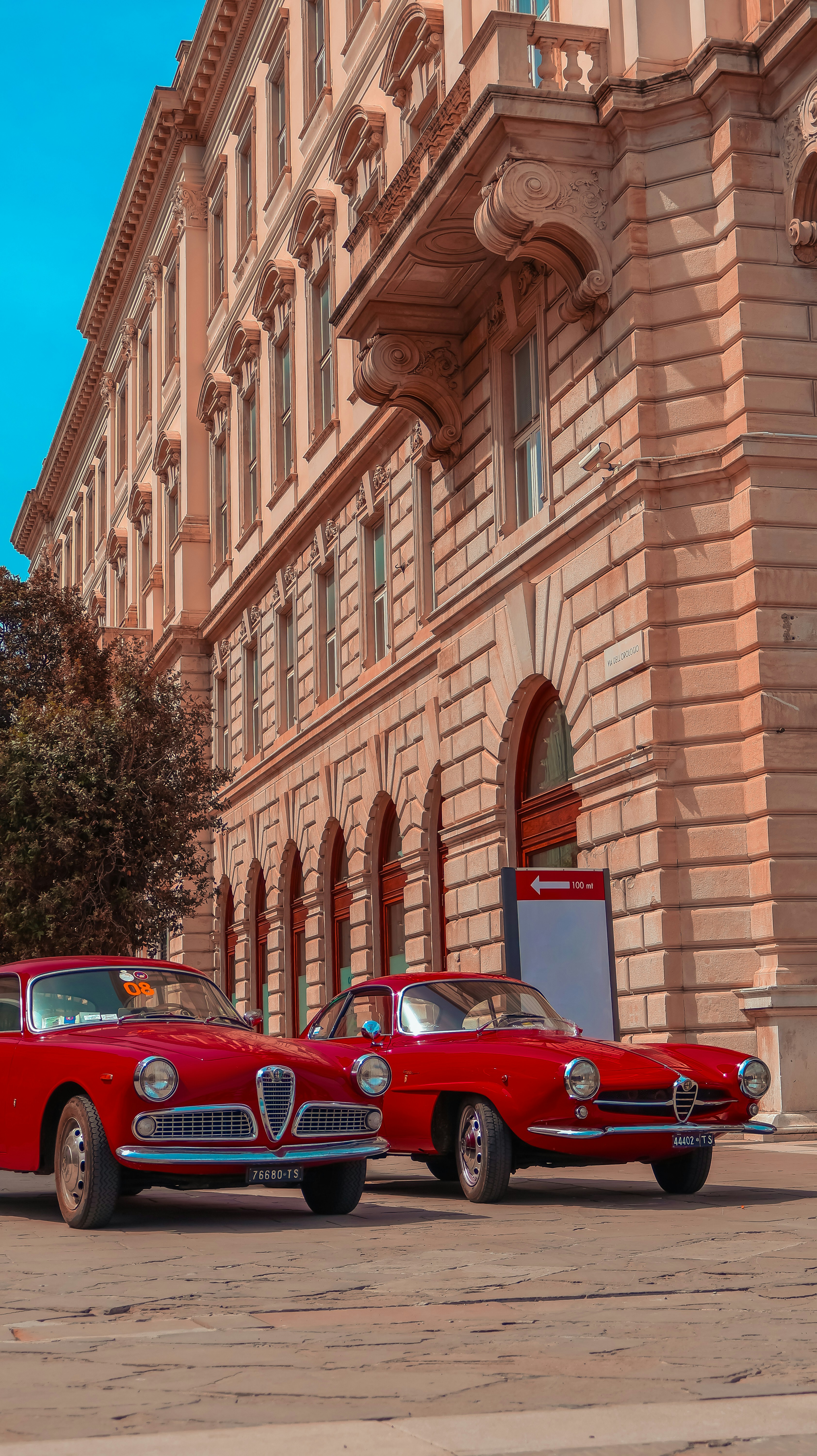 Two classic Alfa Romeo cars parked in front of a historic building with ornate architecture and a clear blue sky.