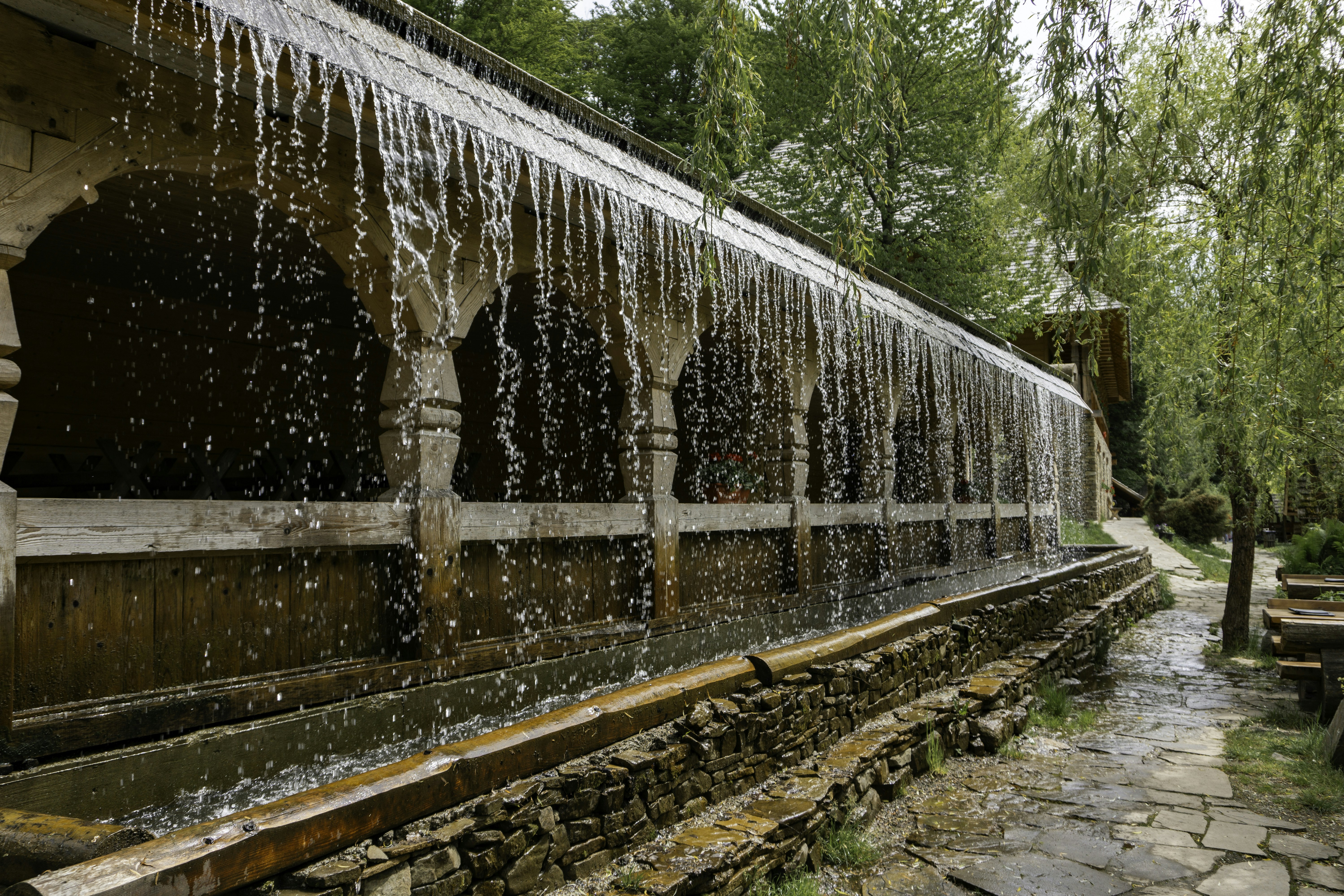 Water cascades over a wooden bridge with stone arches, surrounded by lush greenery.