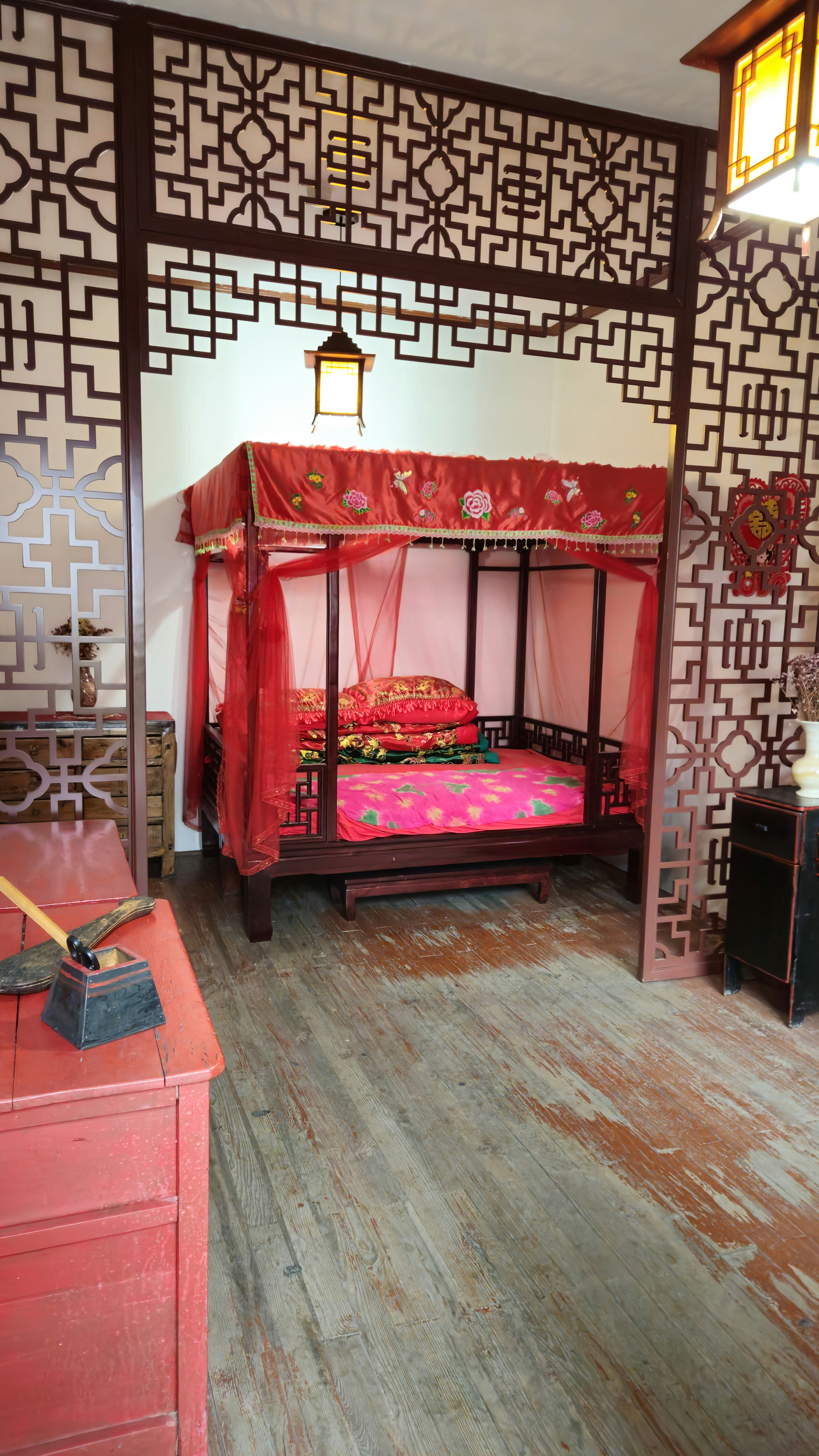 Red canopy bed with floral textiles sits at the center of a room framed by geometric lattice panels. Warm lantern light enhances the heritage atmosphere and highlights the carved wood and textiles.