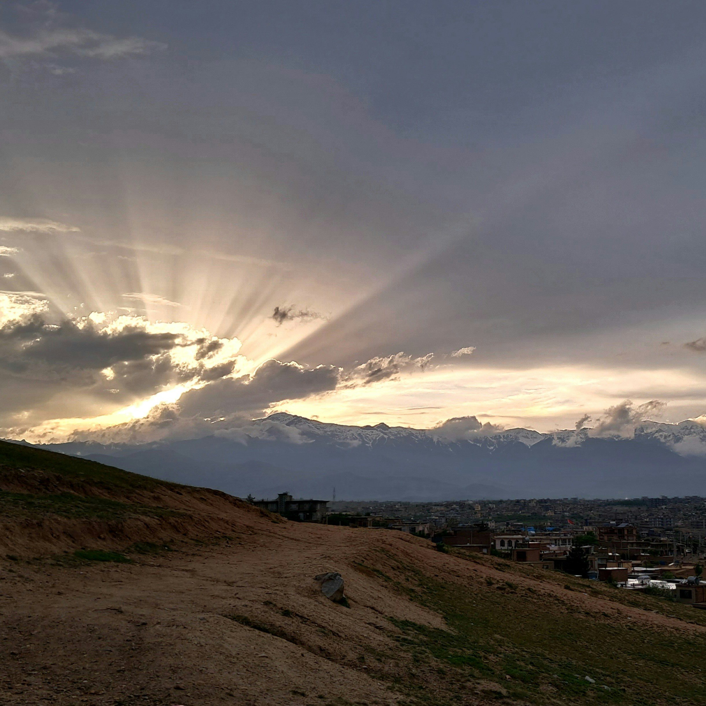 Hillside foreground frames a distant city beneath dramatic sunbeams piercing clouds. Landscape photograph highlighting natural light spilling over the horizon.