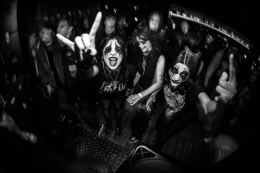 A packed black-and-white concert scene with face-painted fans throwing rock hand signs and leaning toward the camera. The central woman’s open-mouthed expression dominates the frame.