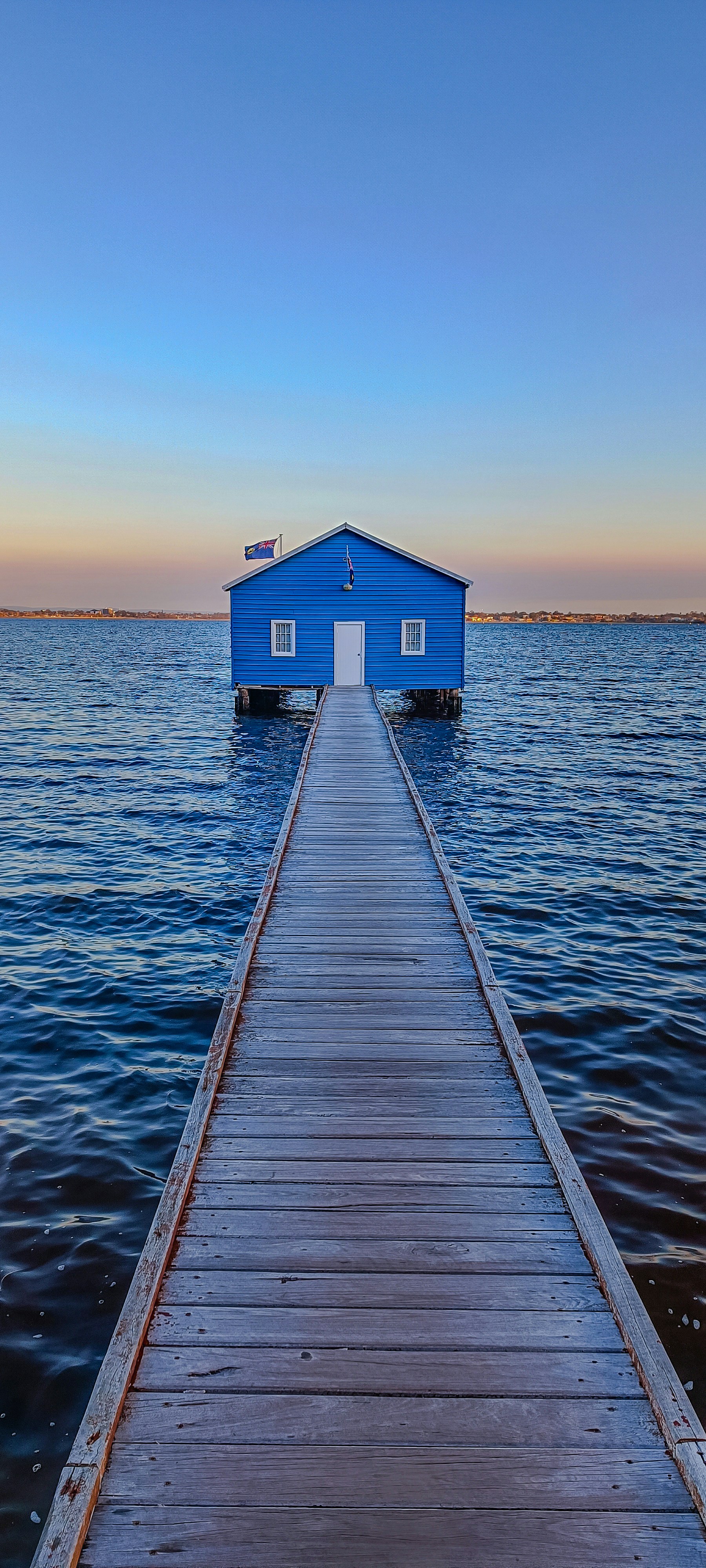A vivid blue boathouse sits at the end of a weathered wooden pier stretching into calm water beneath a warm sunset sky.