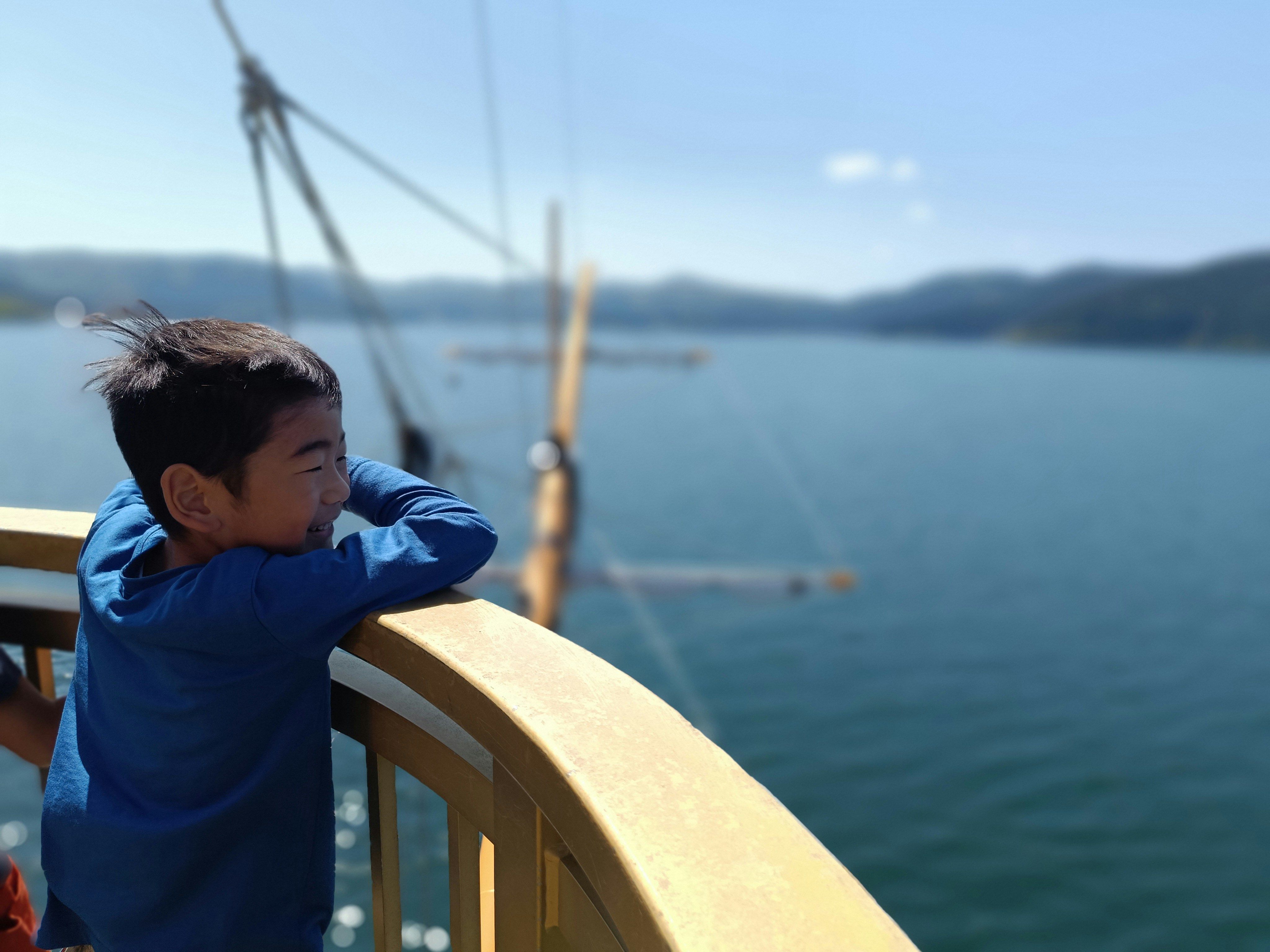 A child in a blue jacket leans on a wooden railing, gazing toward the distant blue water and horizon; the scene is captured on a sunny day near a sailboat. (Photo is a candid capture from a camera phone.)