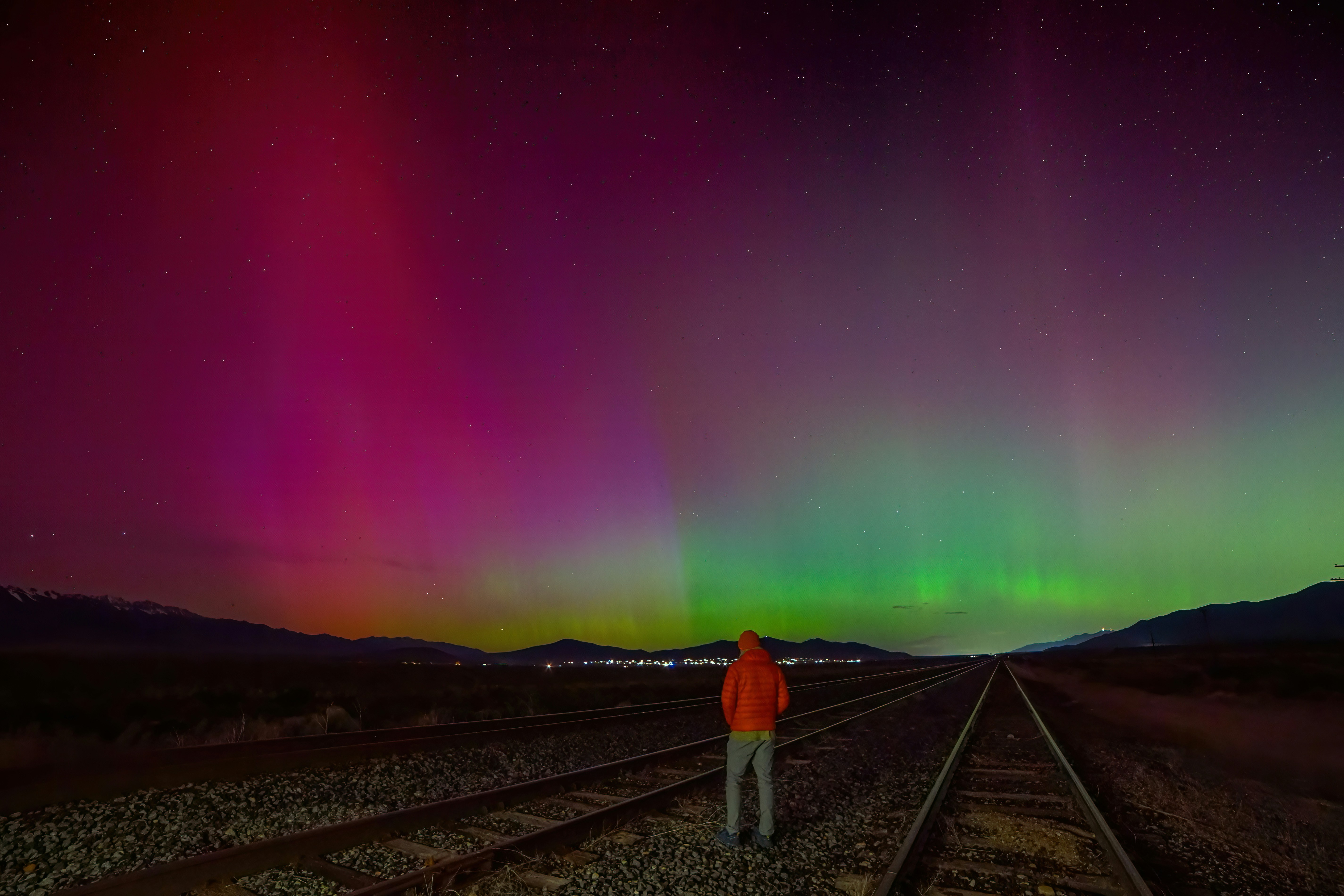 Person standing on railroad tracks under vibrant northern lights in a clear night sky.