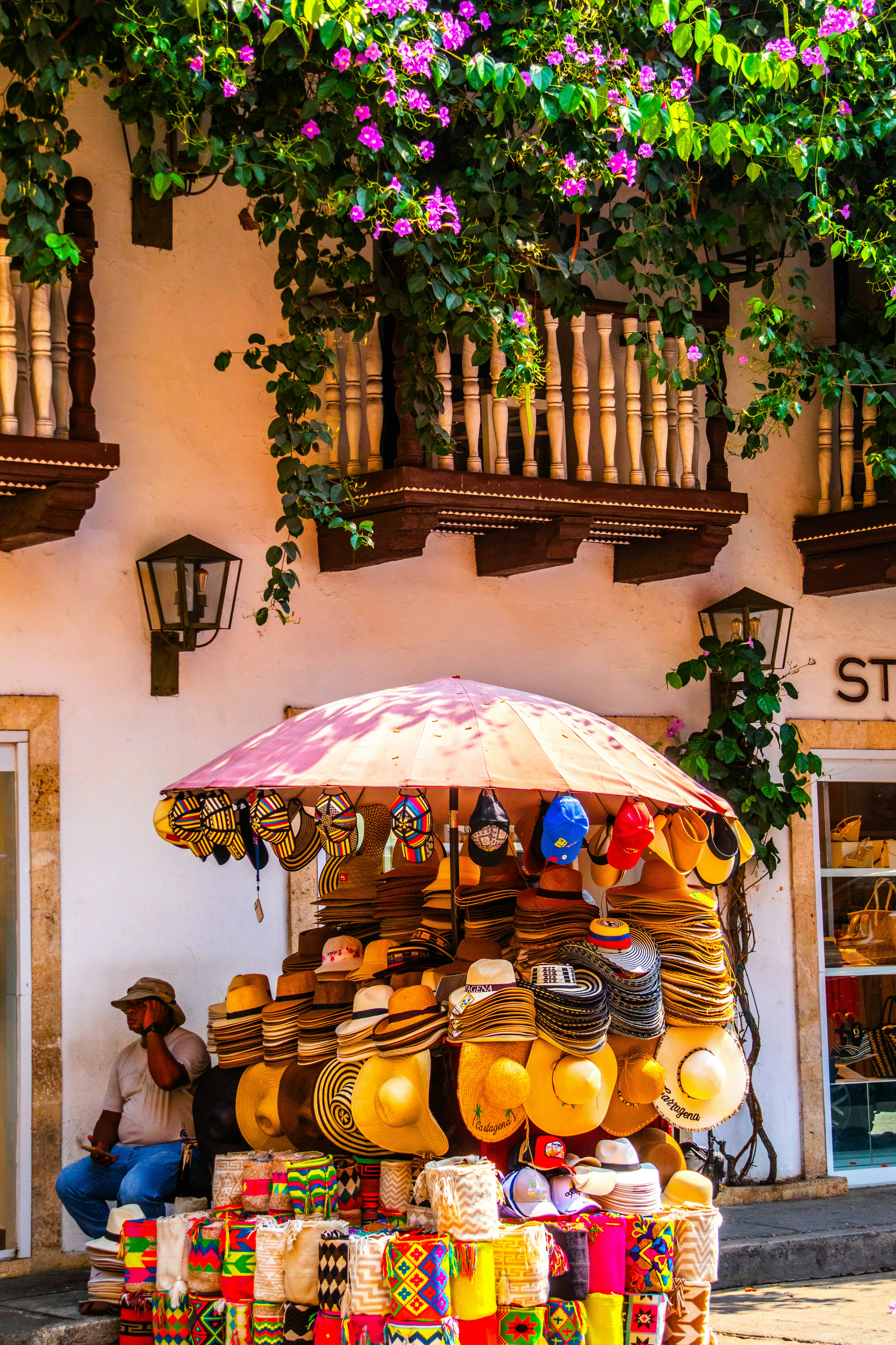 Blush Parasol Over a Kaleidoscopic Hat Stall in Sunlit Street