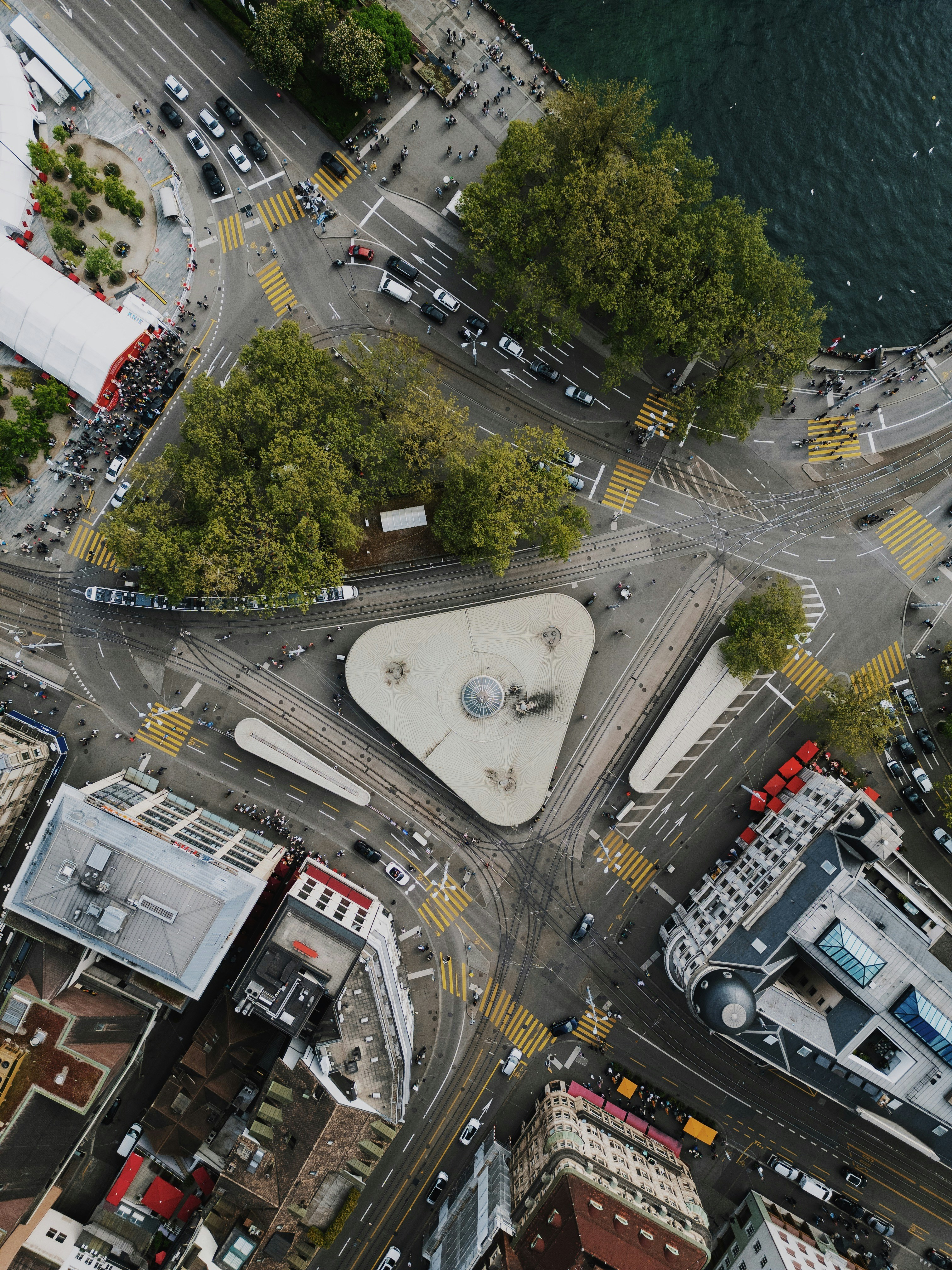 Aerial view of a busy intersection surrounded by trees and buildings, showcasing the intricate layout of urban life. The central fountain serves as a focal point amidst the bustling streets.