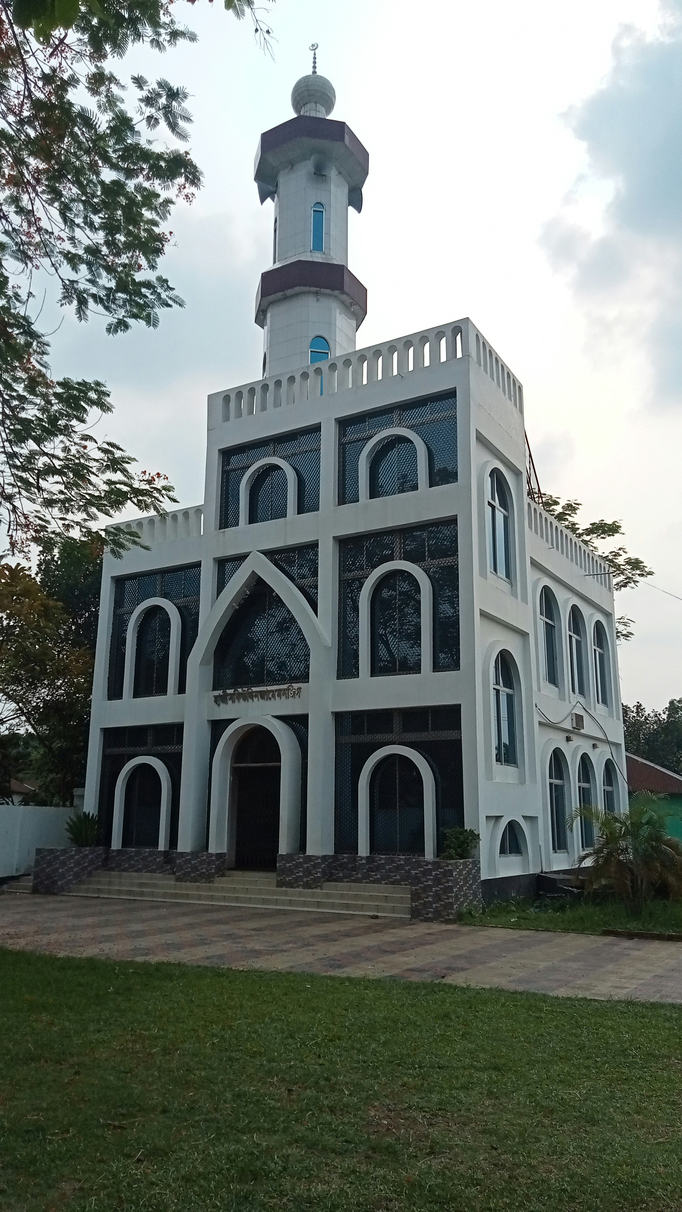 A white, multi-arched mosque façade with a tall minaret rises above a stepped entrance, framed by trees and a manicured lawn.