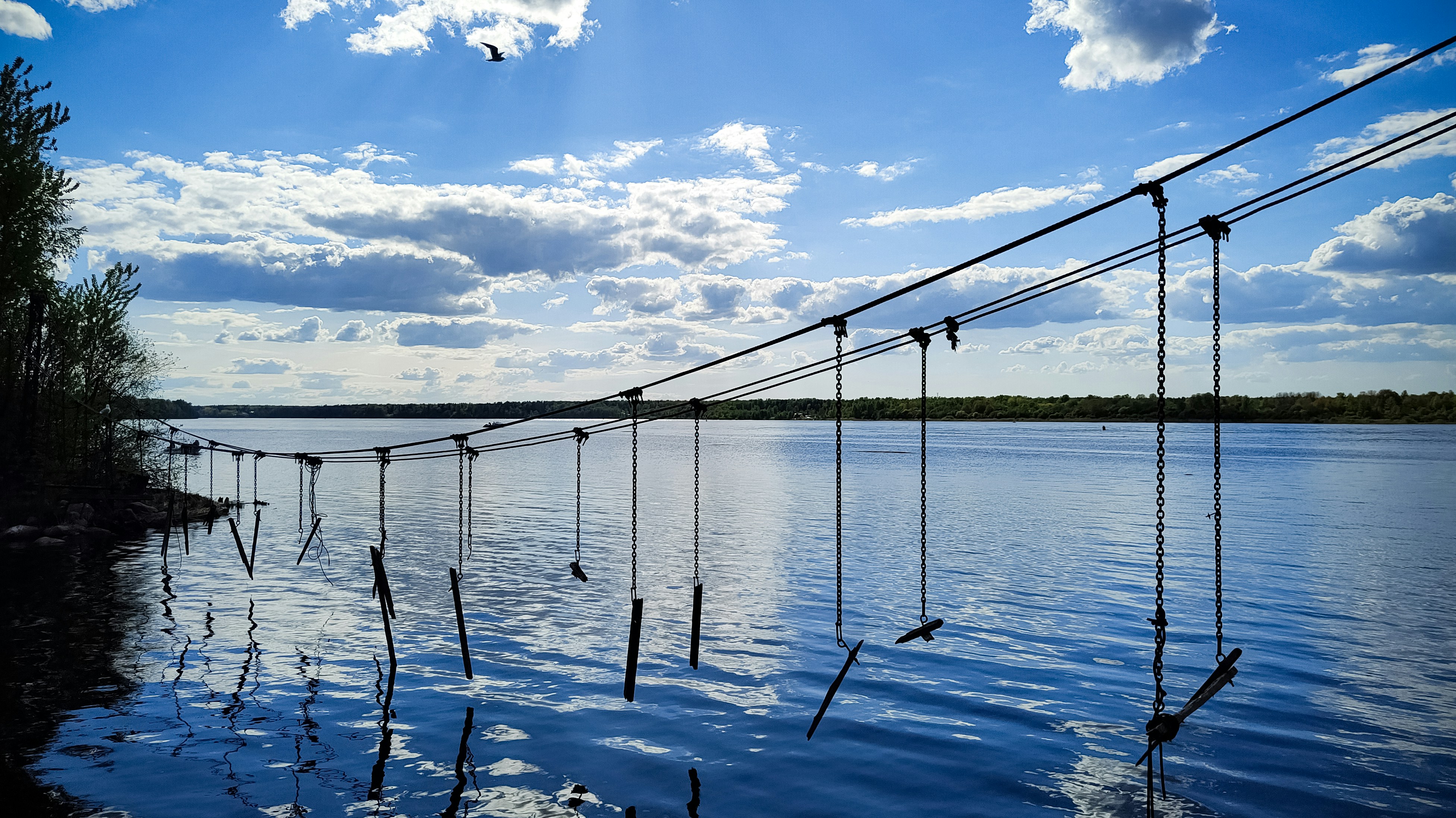 Calm river scene with a suspended rope bridge and vertical posts, reflecting a sky with scattered clouds. The composition highlights depth and the bridge's repeating lines against the water.