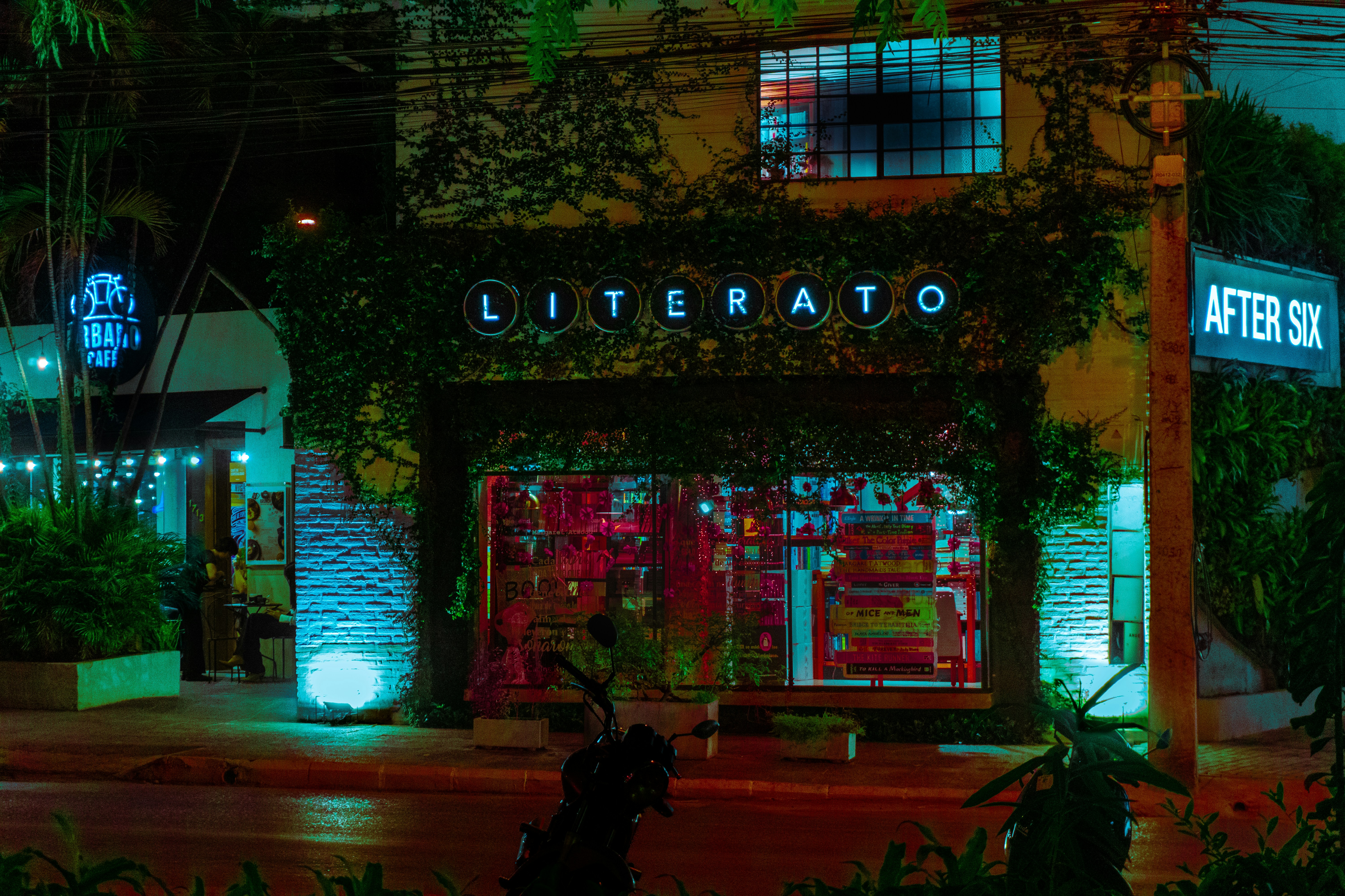 Motorcycle parked in front of a brightly lit building adorned with vibrant neon signs and lush greenery at night.