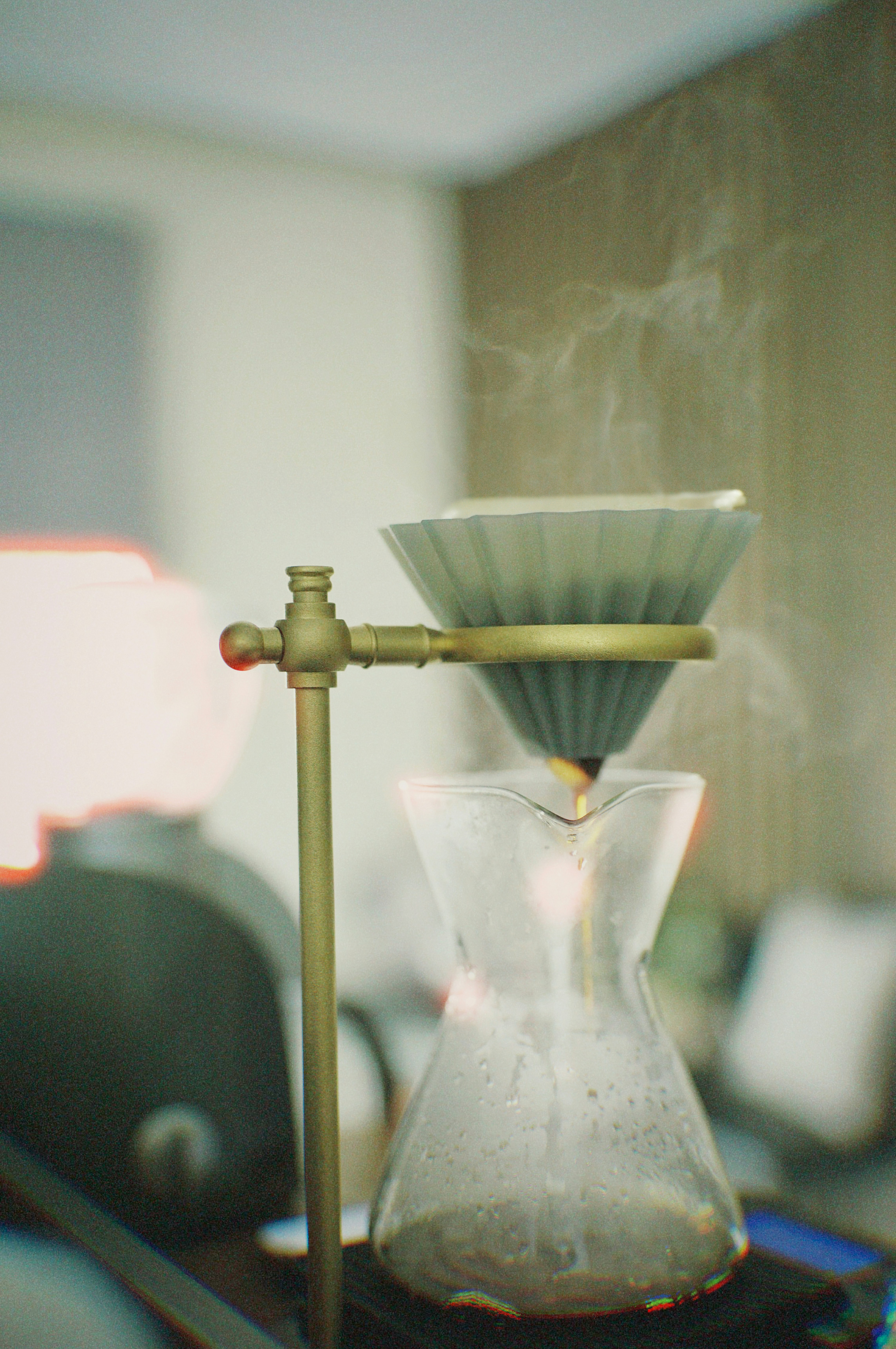 A brass pour-over stand supports a ceramic conical dripper perched above a glass carafe in a softly lit room.