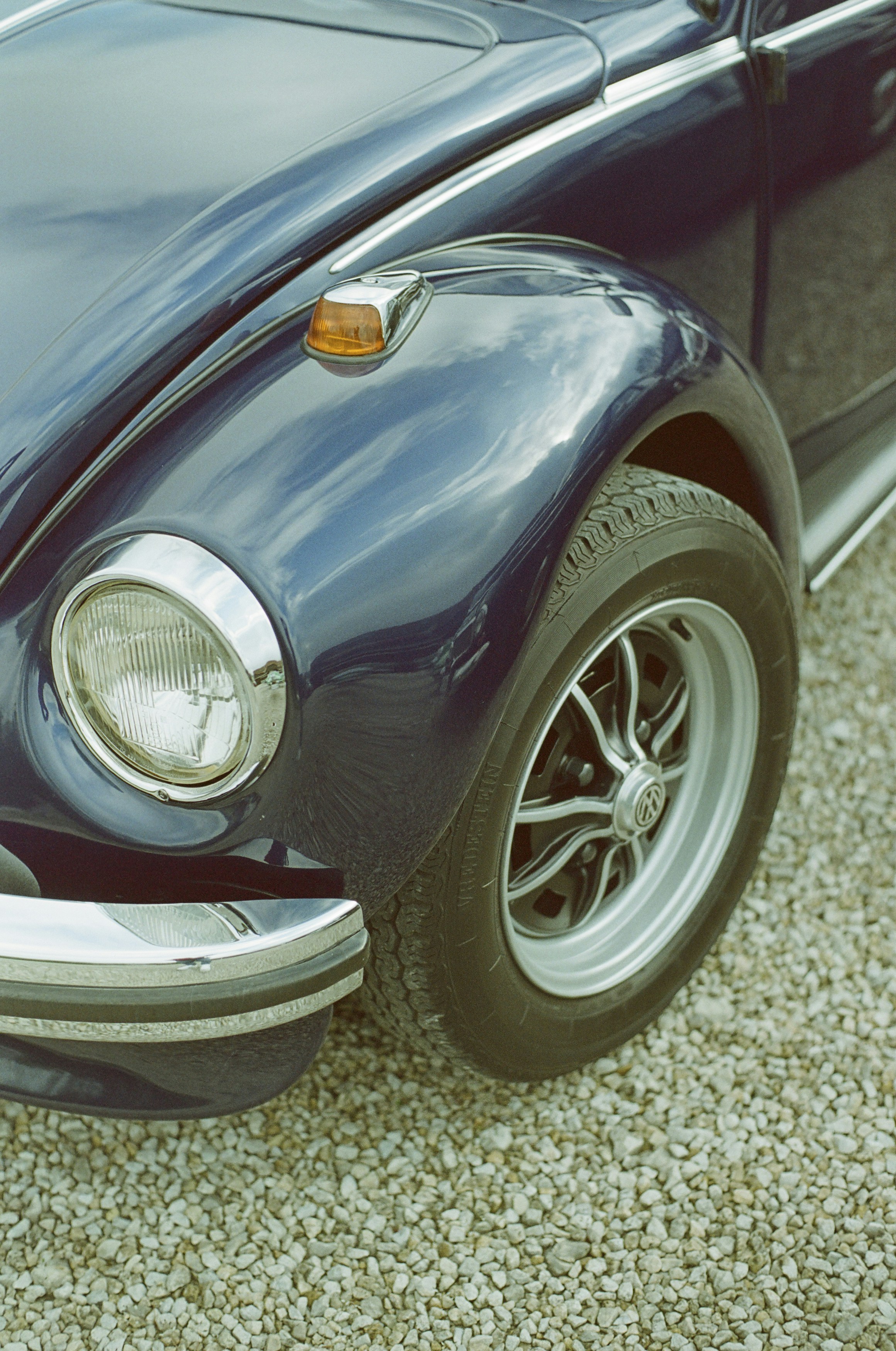 Close-up of a classic blue VW Beetle's front end on gravel, highlighting the curved body and intricate wheel design.