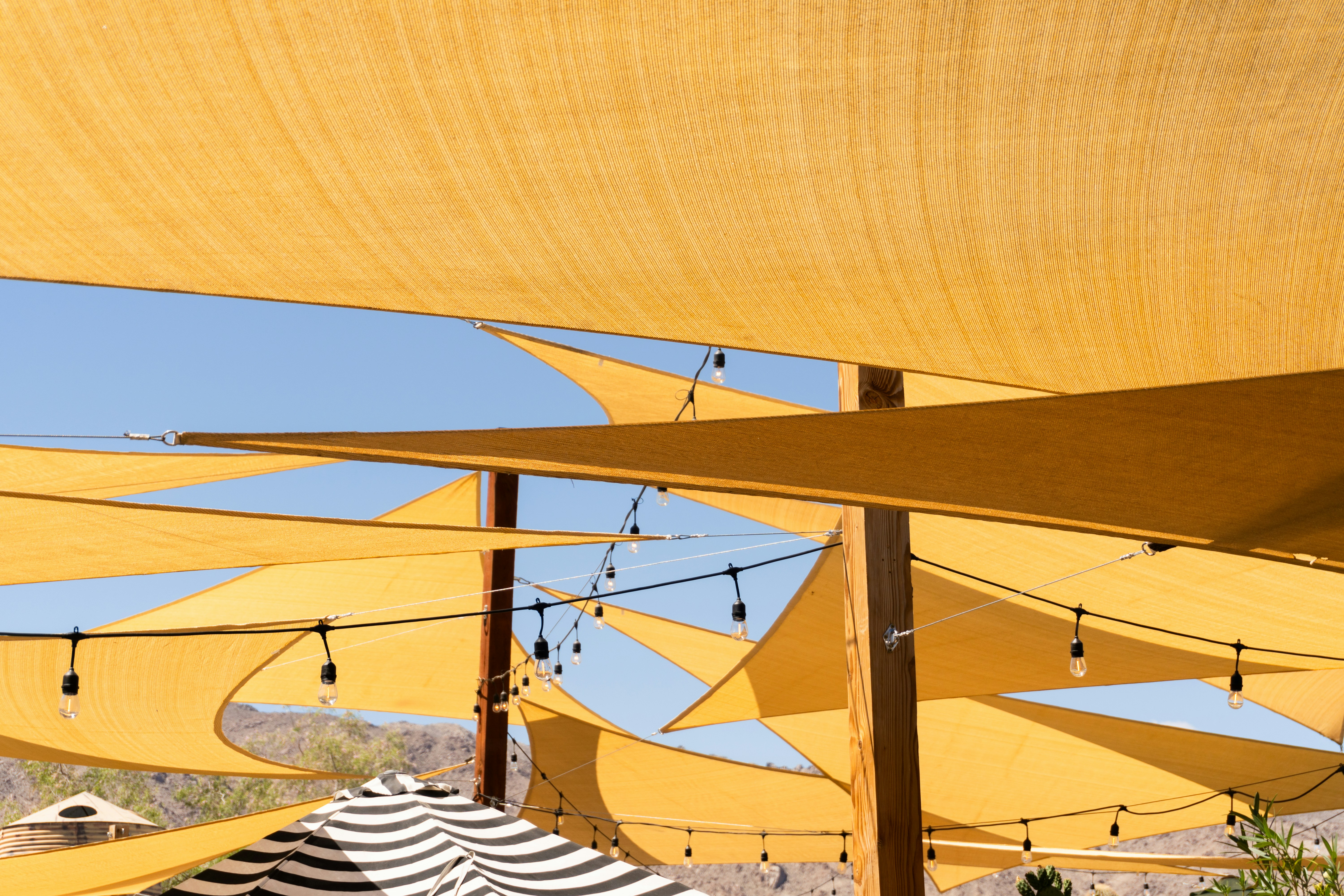 Yellow sail-like shades stretch across a sunny outdoor space with string lights and a striped umbrella.