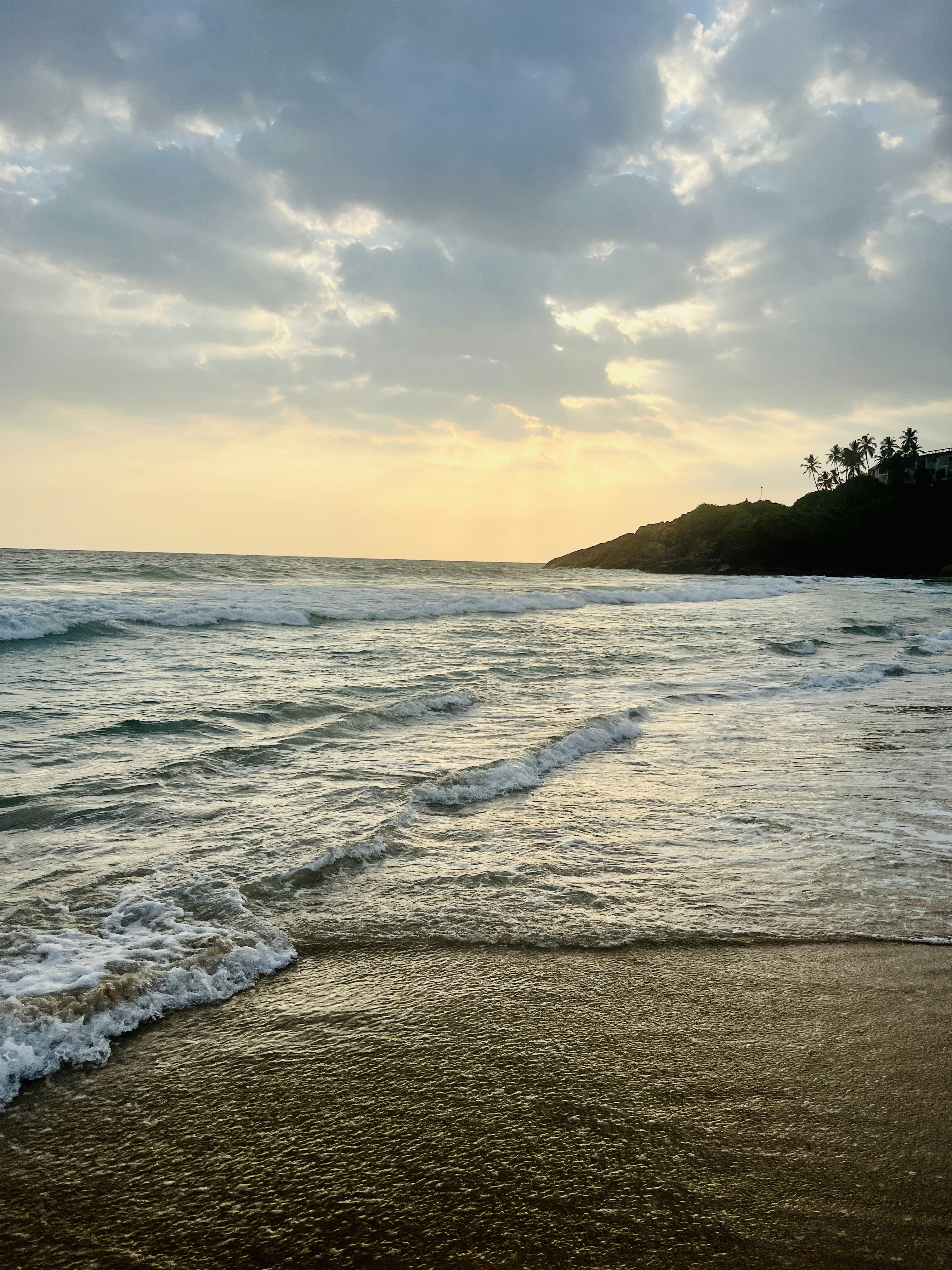 Gentle waves lap against the shore under a cloudy sky, with a distant silhouette of palm trees on a hillside. The soft light of dusk casts a serene atmosphere.