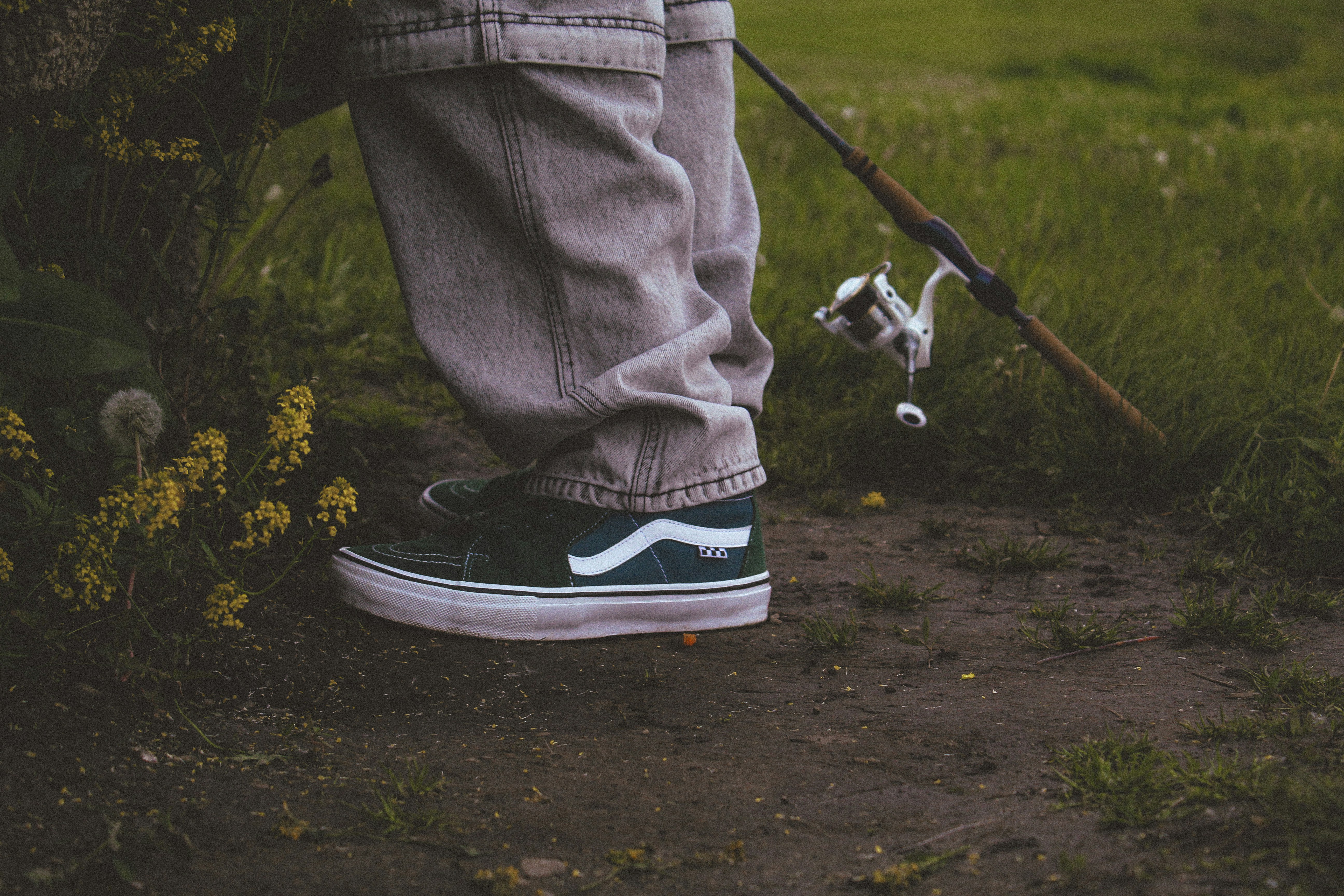 A person stands on a dirt path, wearing green Vans sneakers, with fishing gear in the background amidst a lush, grassy landscape.