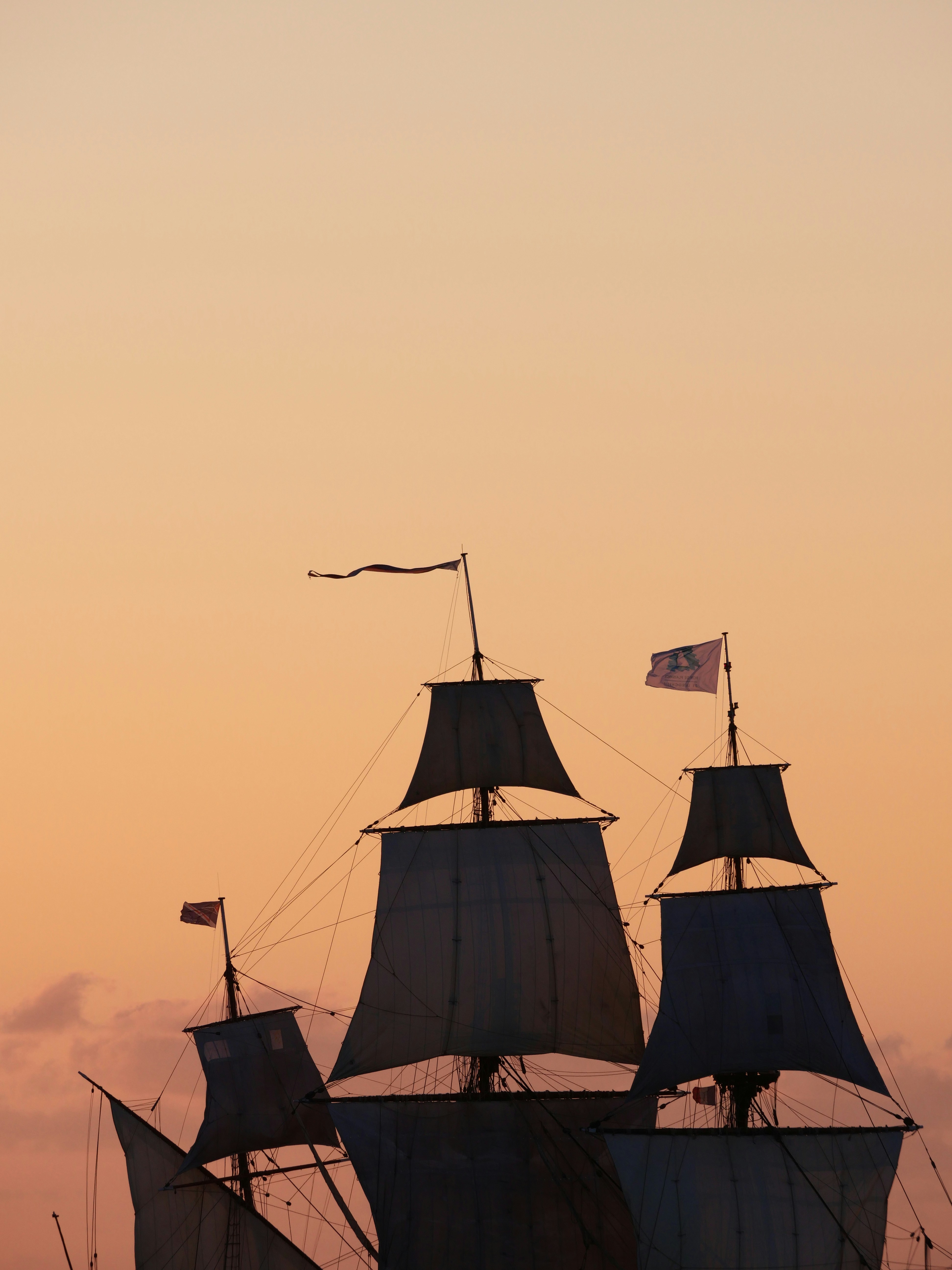 Silhouetted three-masted schooner sails rise against a peach-orange sunset, creating a calm maritime silhouette.