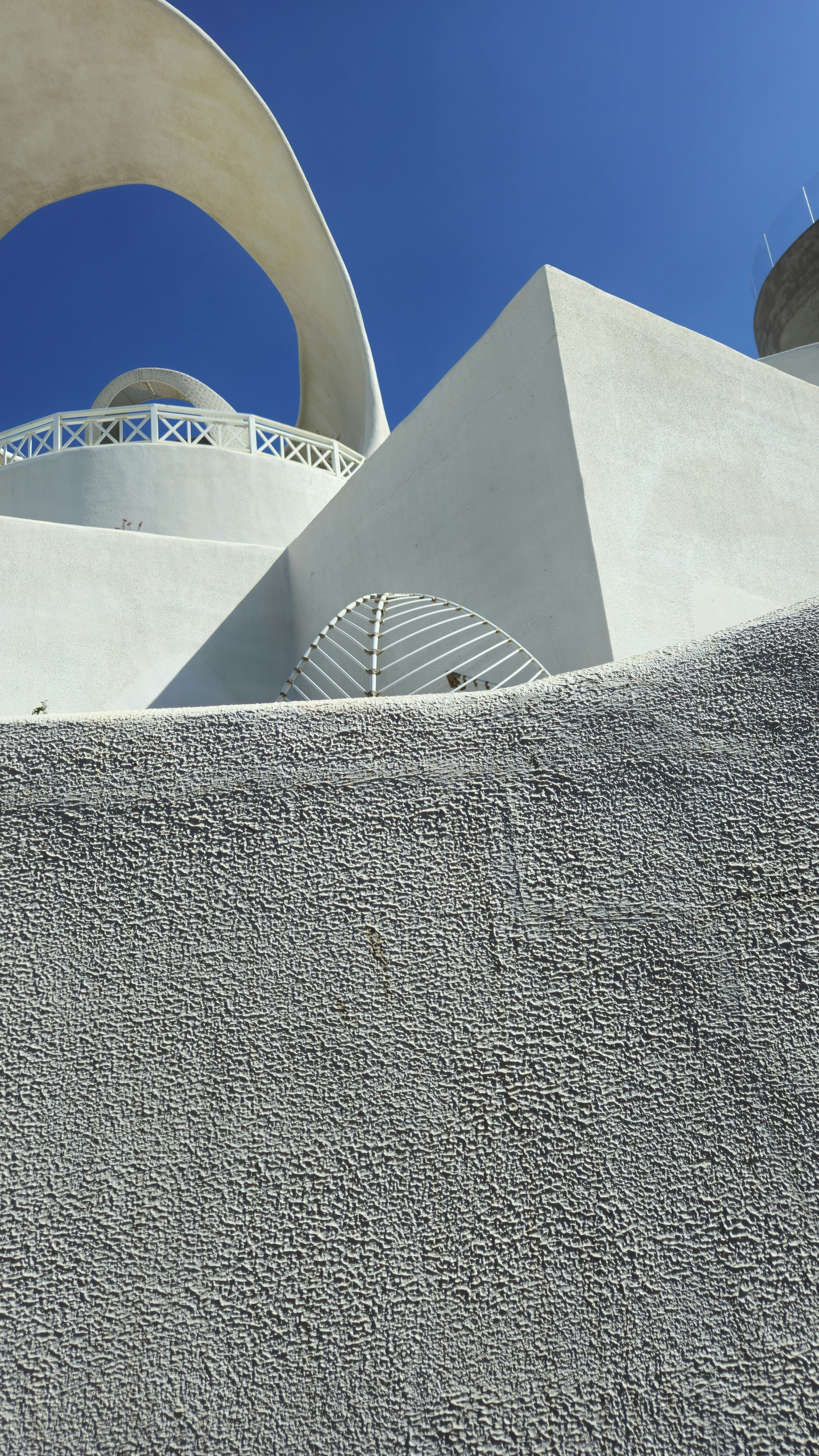 White modernist architecture with a sweeping arch and angular planes set against a clear blue sky, foregrounded by a textured stucco wall.