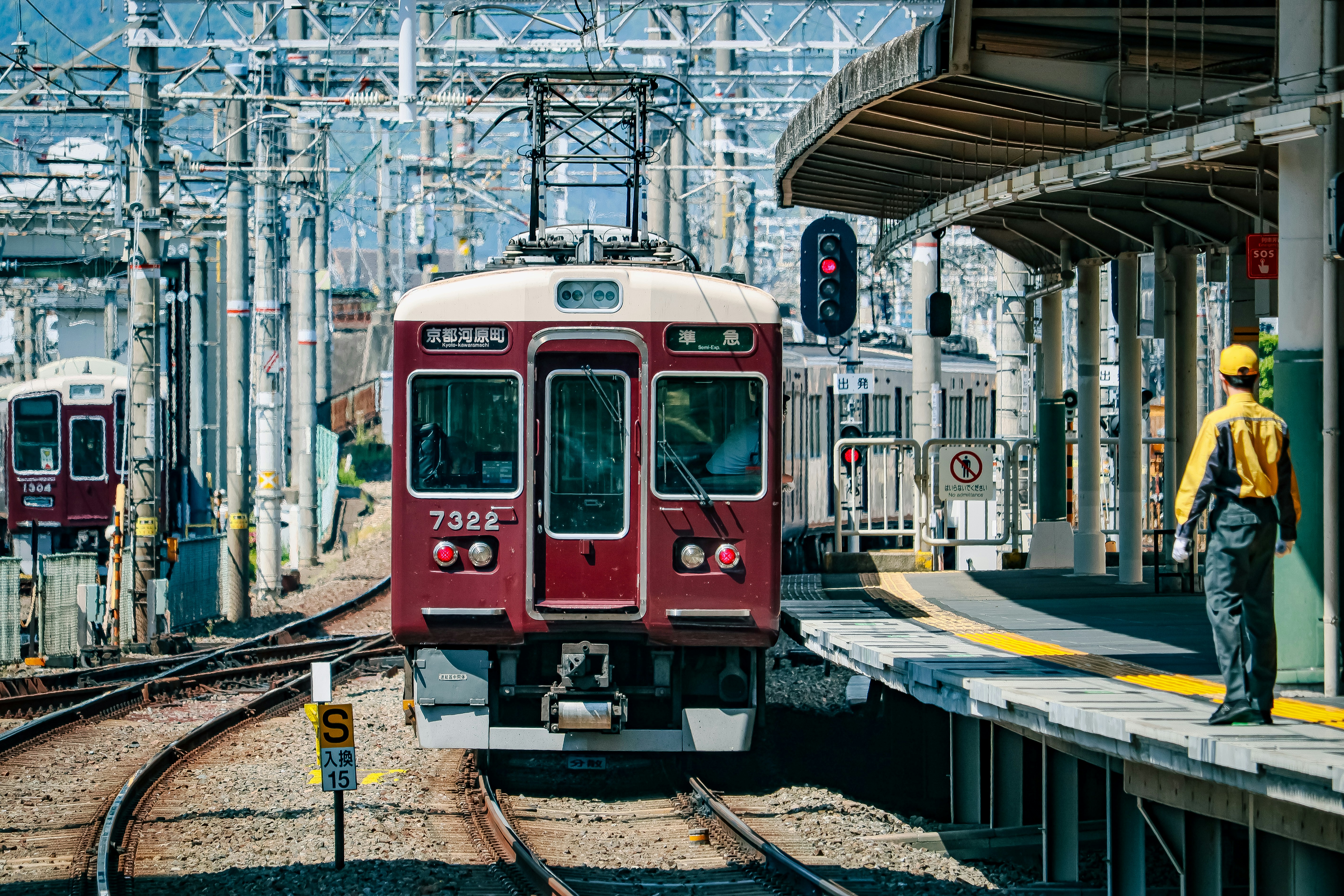 Red train approaching a station platform with a person standing nearby and overhead wires visible.