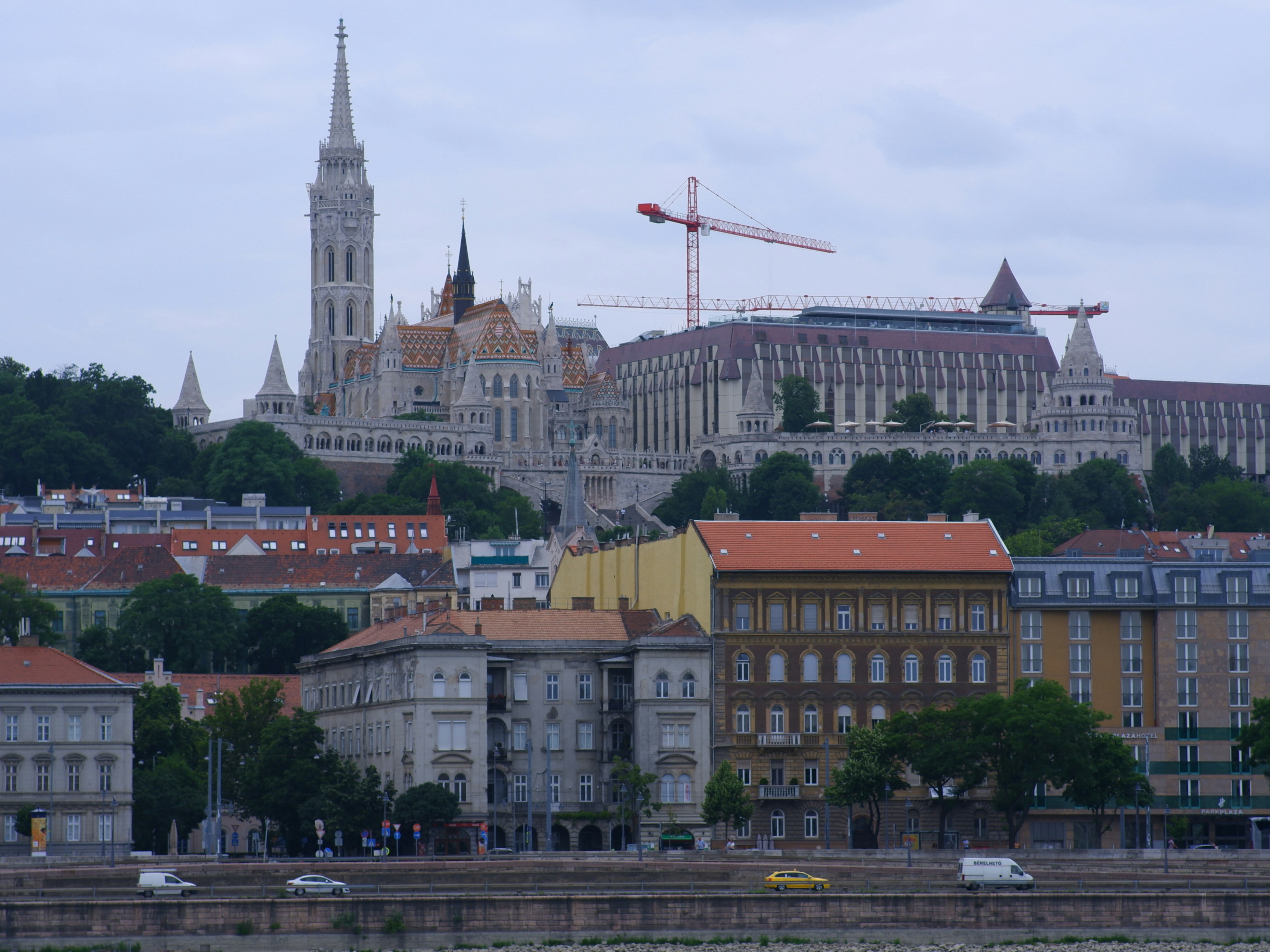 A daytime cityscape of Buda Castle hill centered on a tall spire with a crane crossing the skyline. Colorful riverfront buildings frame the foreground.