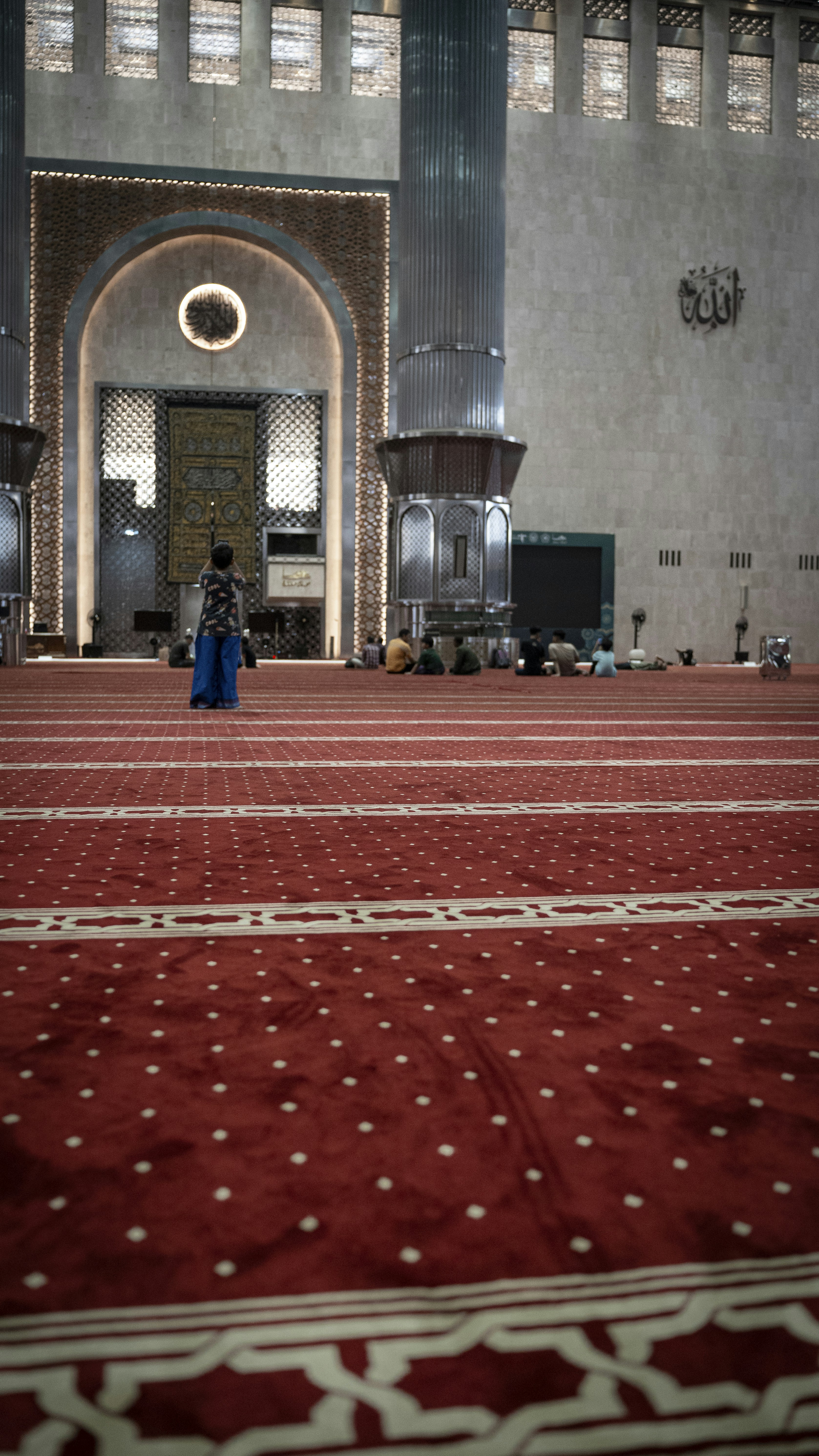 A child stands in a grand mosque, gazing towards the intricately designed entrance, while worshippers are seated in the background. The rich red carpet enhances the serene atmosphere.
