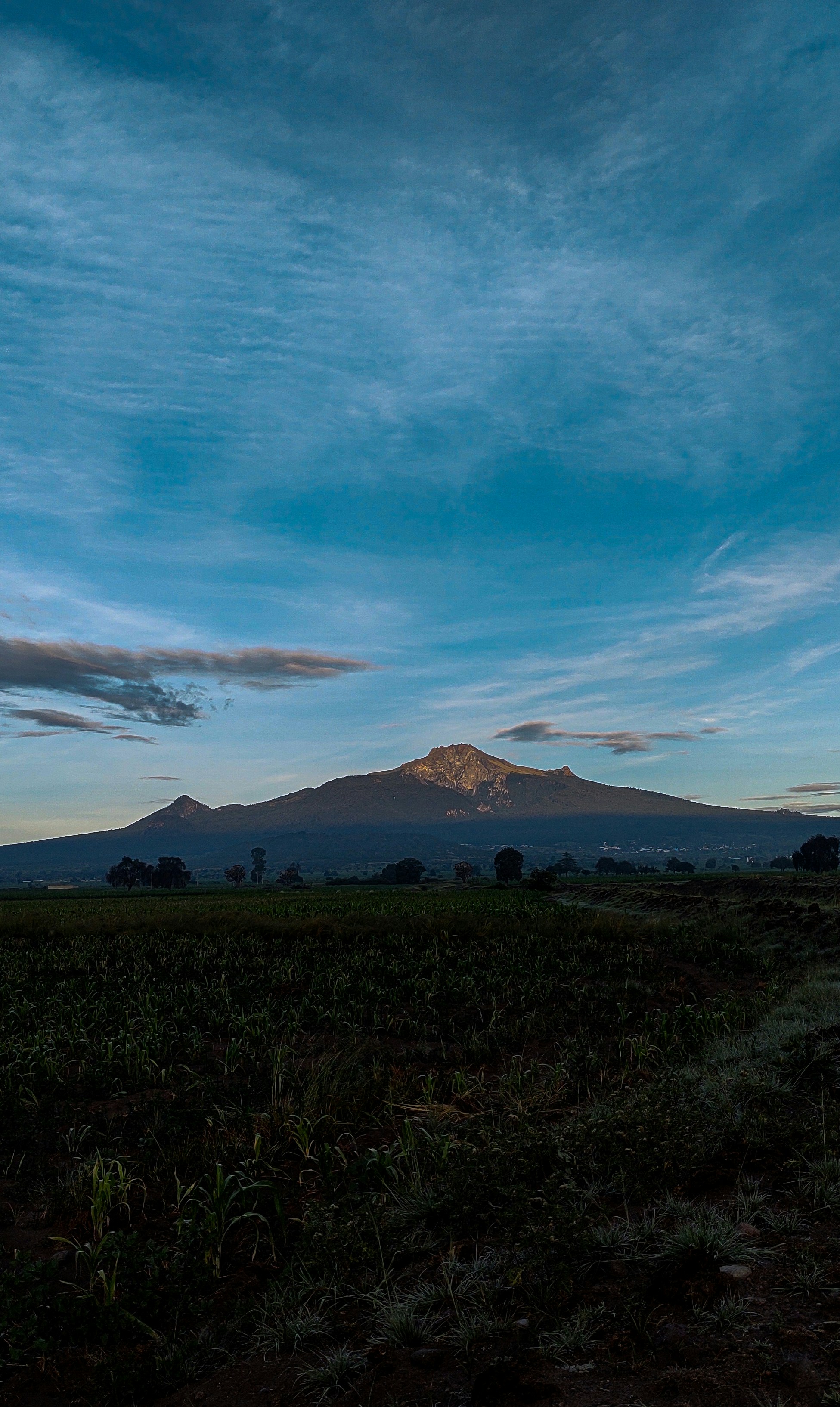 Landscape photograph of a mountain ridge rising above a dark foreground field under a broad blue sky with wispy clouds.