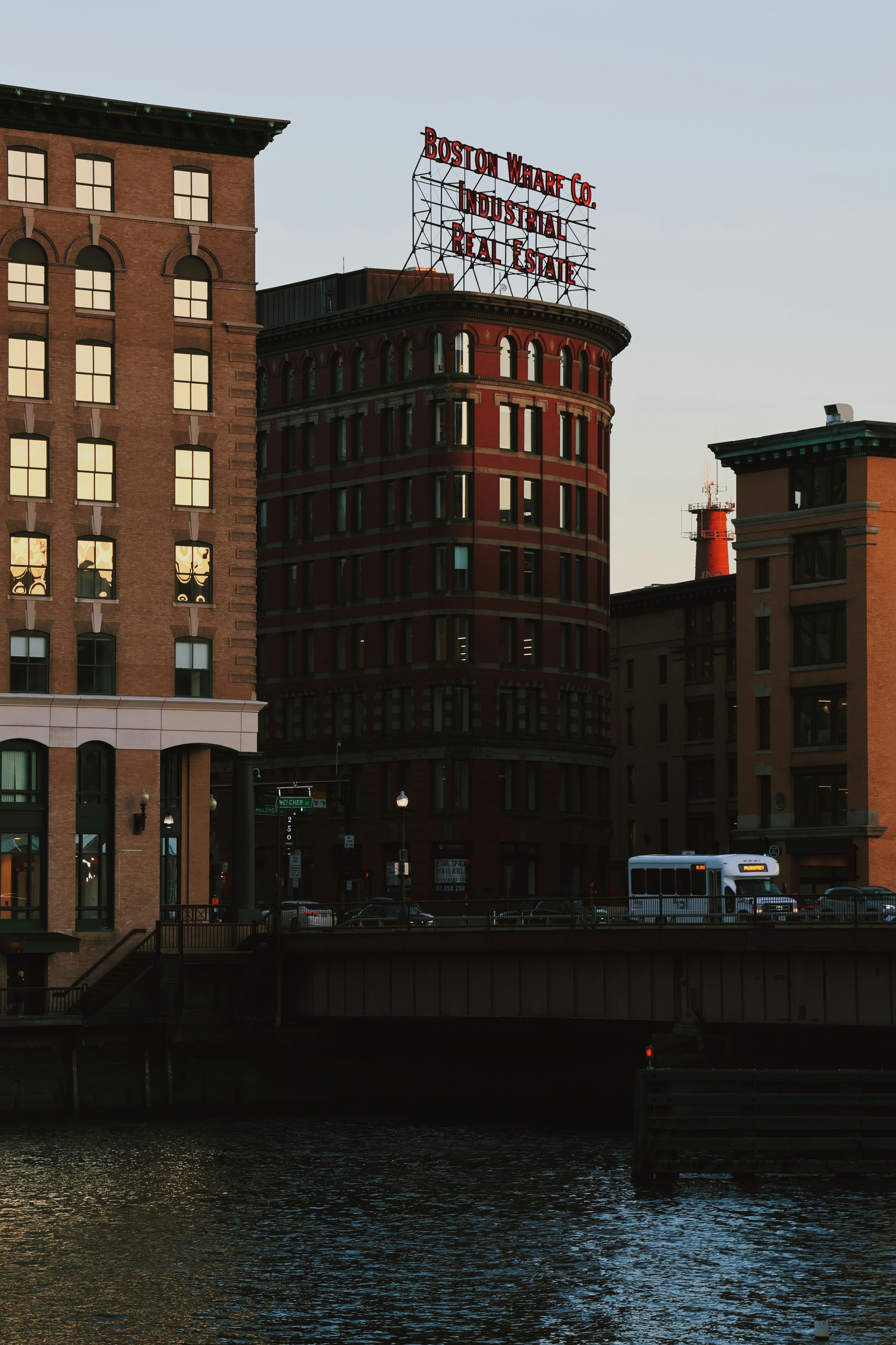 Cityscape photograph of a rounded brick building with a vintage rooftop sign, flanked by other brick towers along a waterfront.