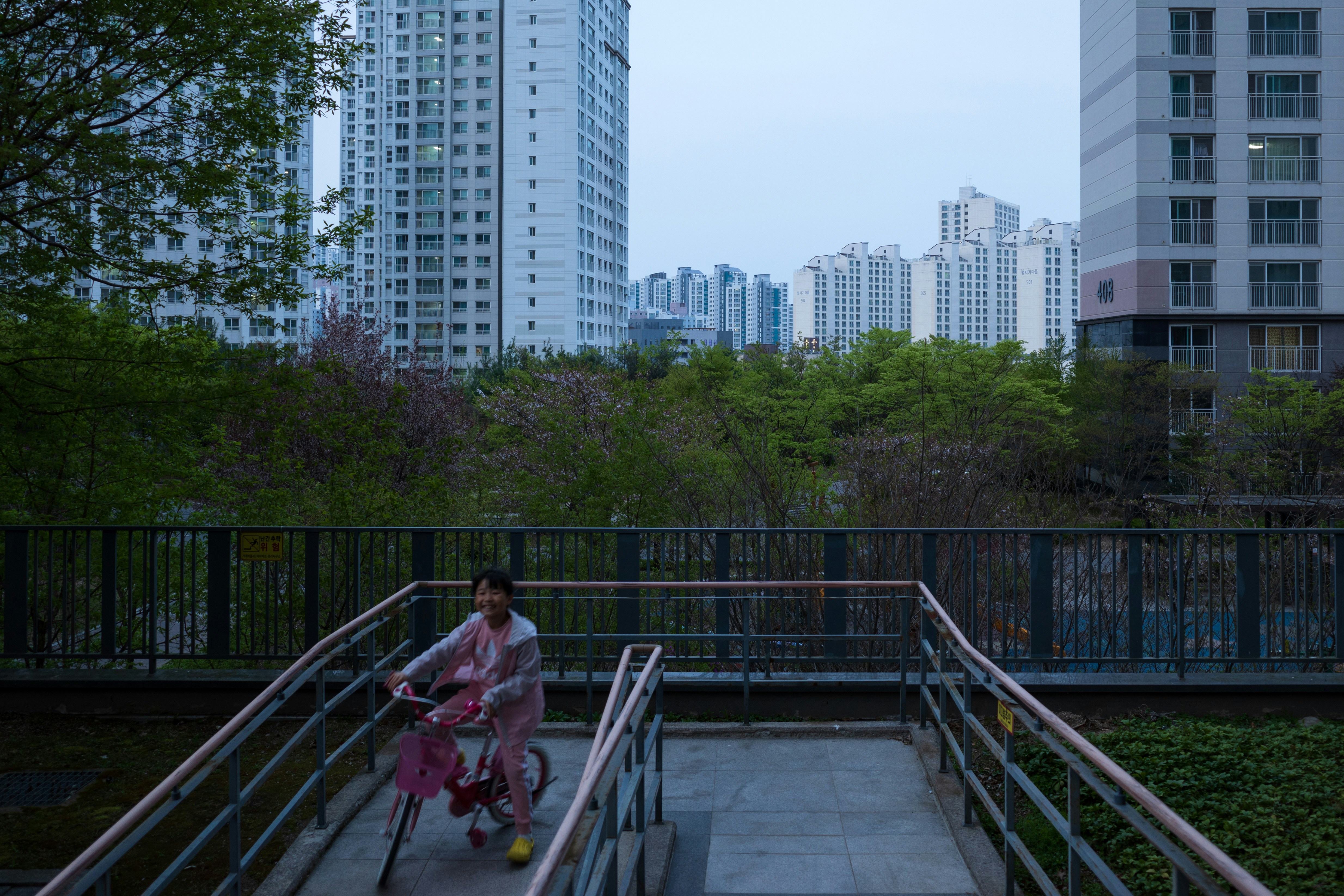 Child riding a pink bicycle down a ramp with high-rise buildings and greenery in the background.