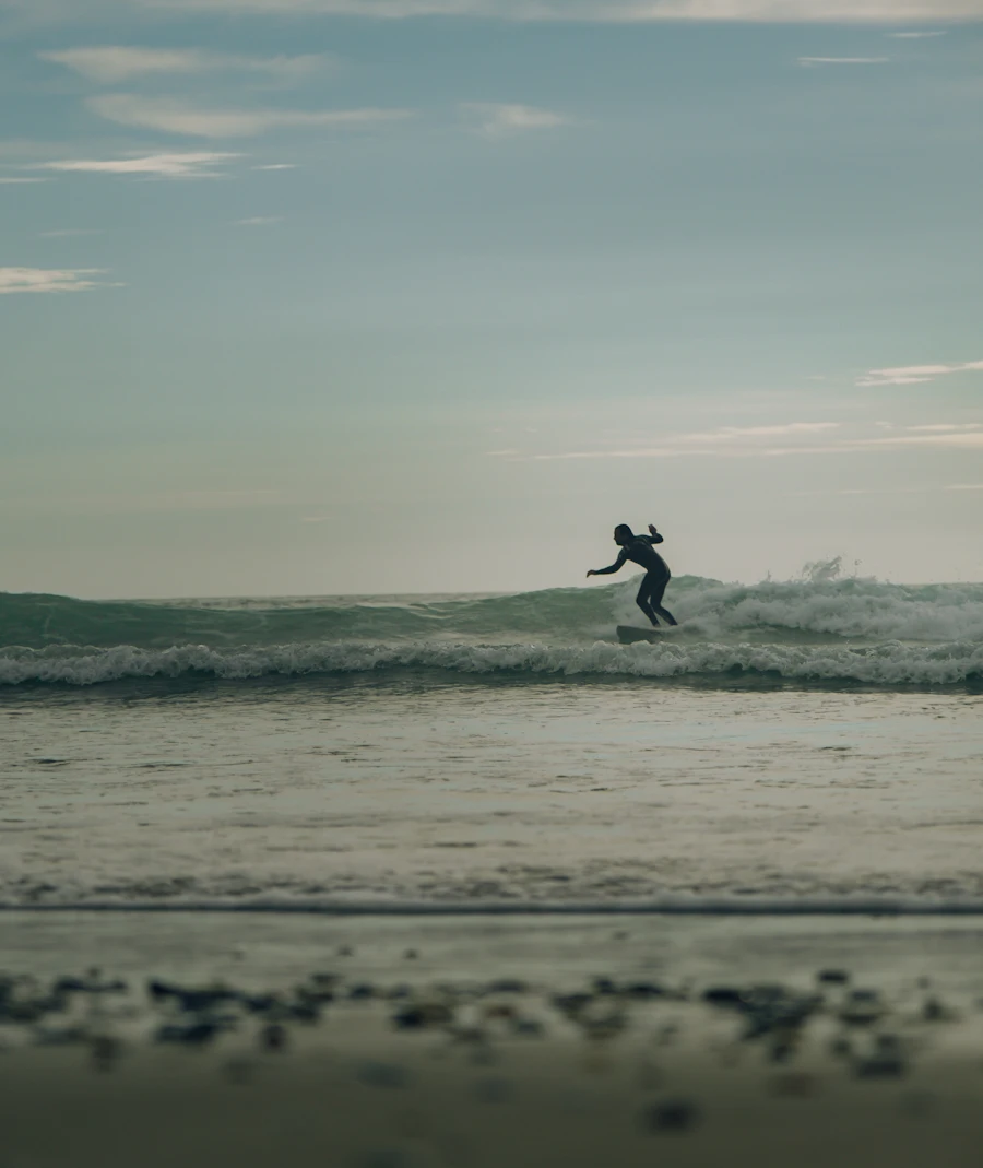 A lone surfer glides along a small breaking wave near a windswept beach at dawn.
