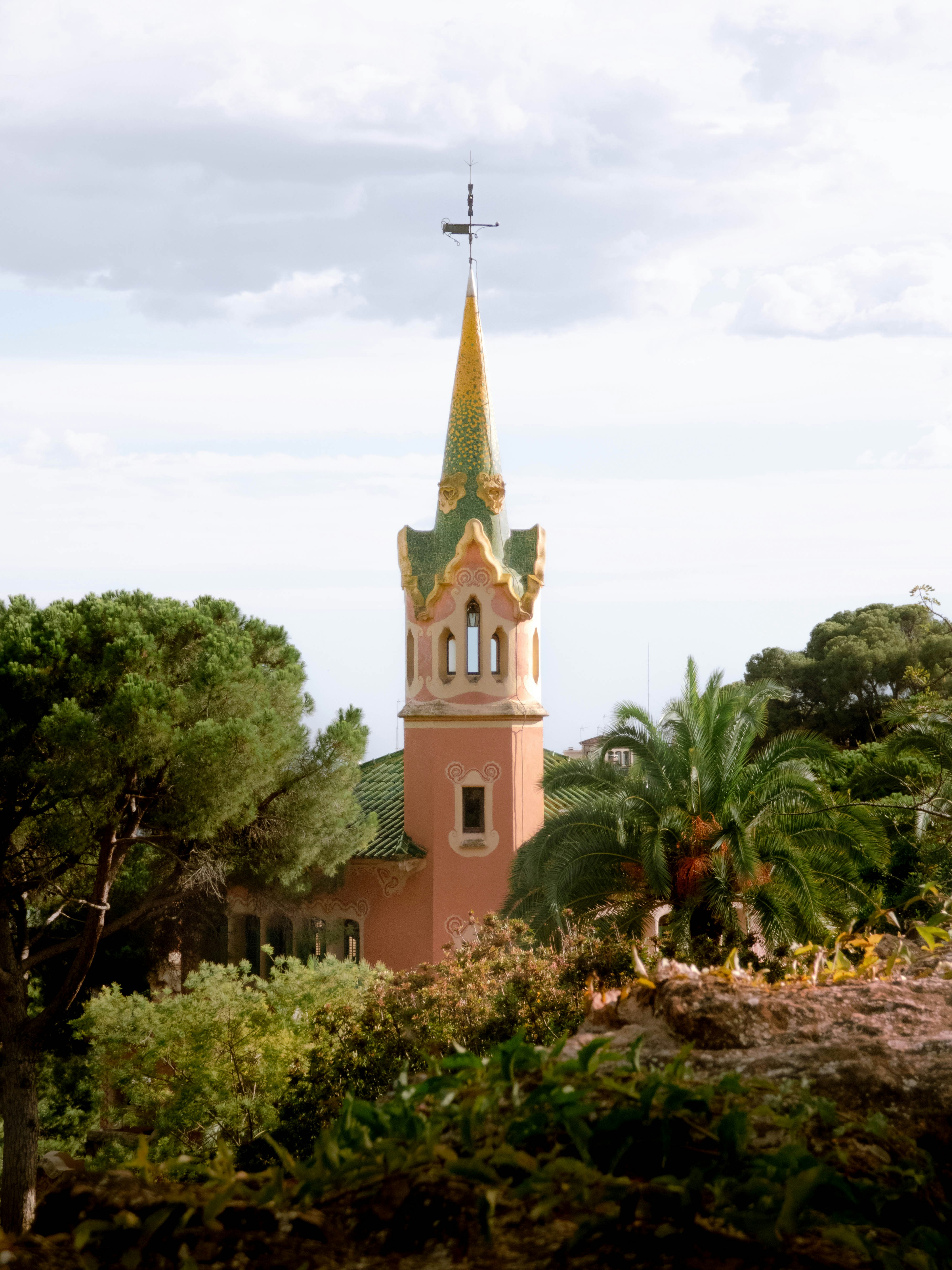 Ornate church steeple with a cross surrounded by lush greenery in Park Güell, Barcelona.
