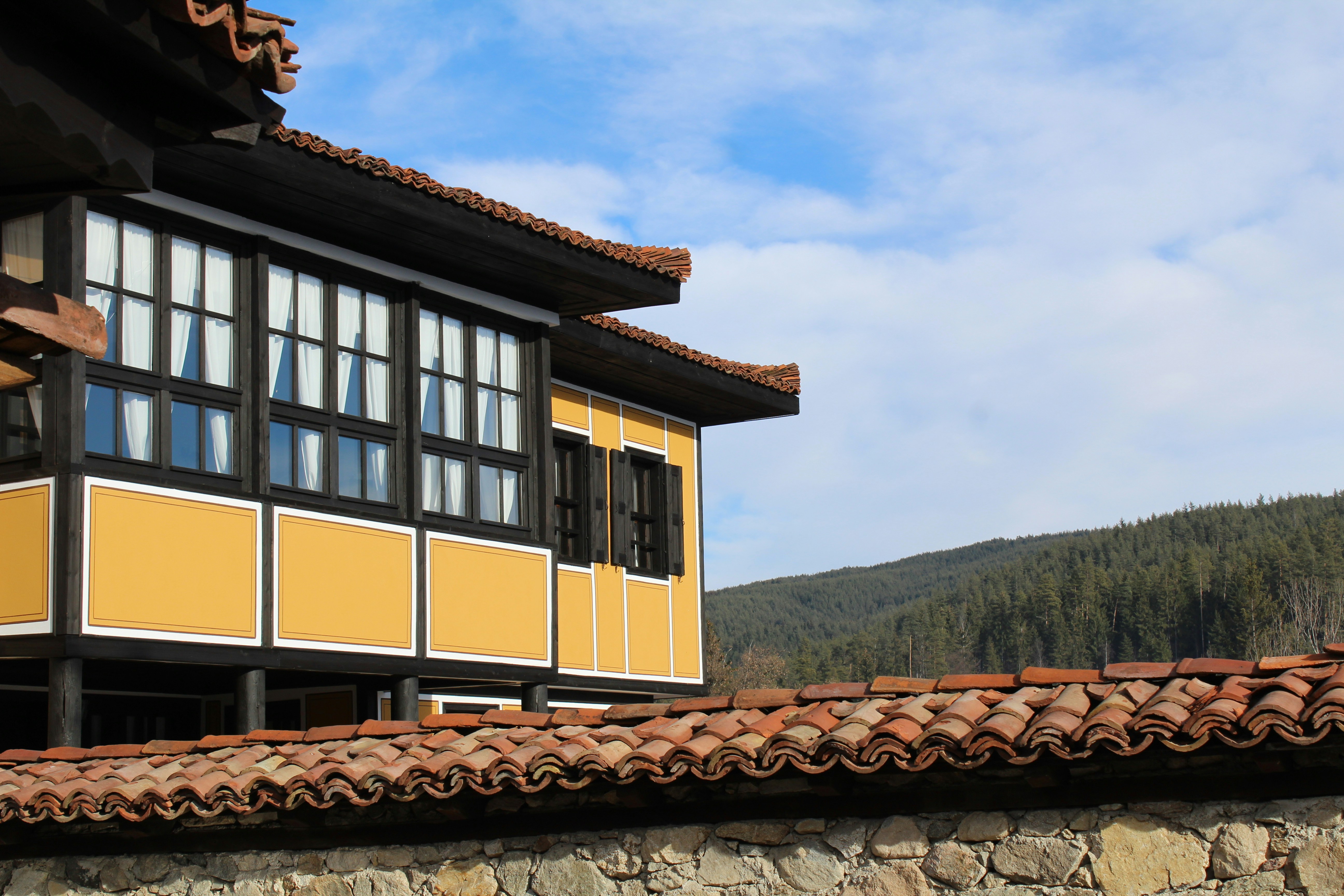 Yellow and black wooden house with large windows against a backdrop of rolling hills and a blue sky. The traditional roof tiles add a rustic charm.