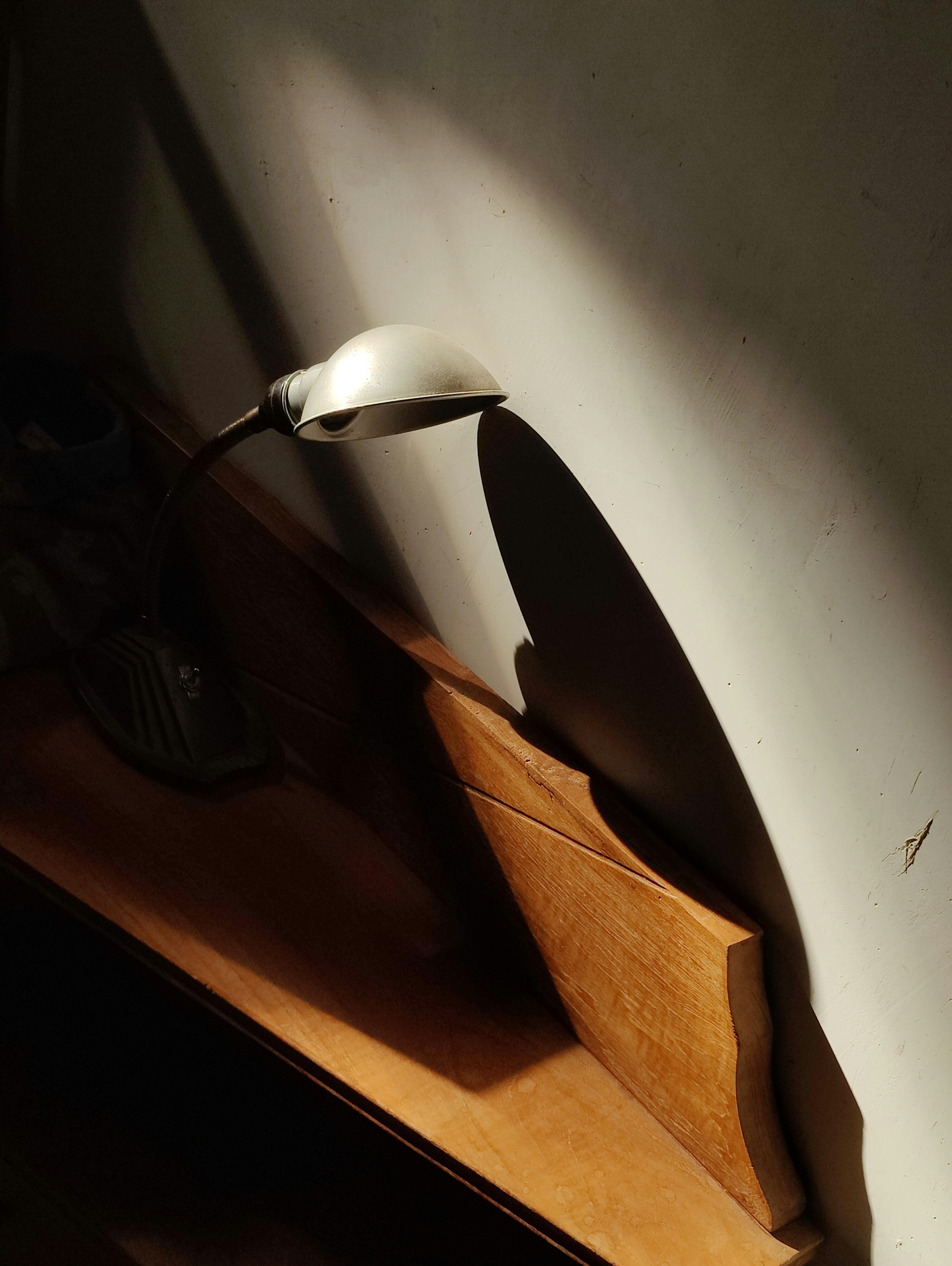 Dimly lit photograph shows a small desk lamp casting a diagonal beam onto a curved wall above a wooden shelf.
