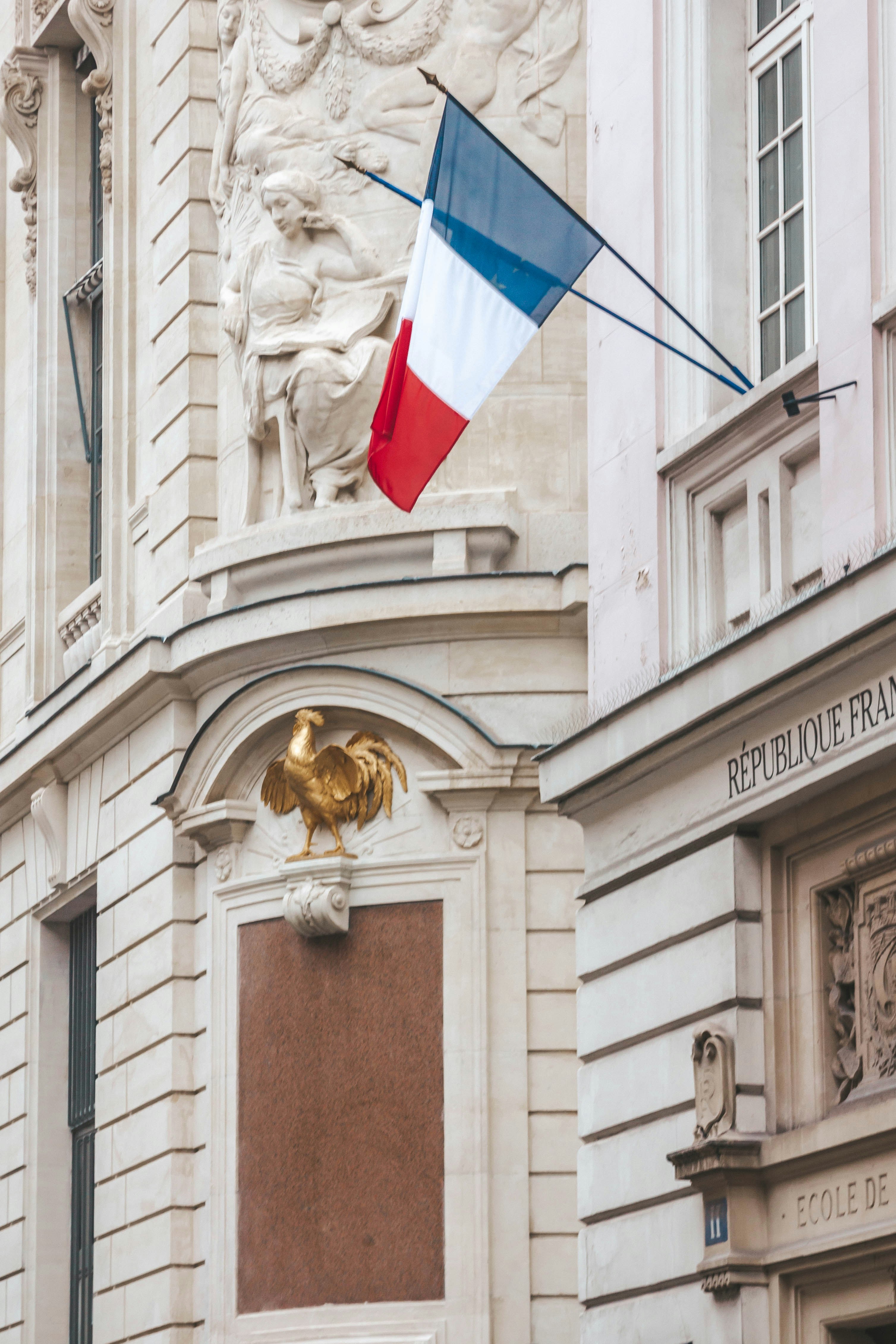 French flag fluttering beside a golden rooster emblem on a historic building facade, showcasing national heritage and artistry.
