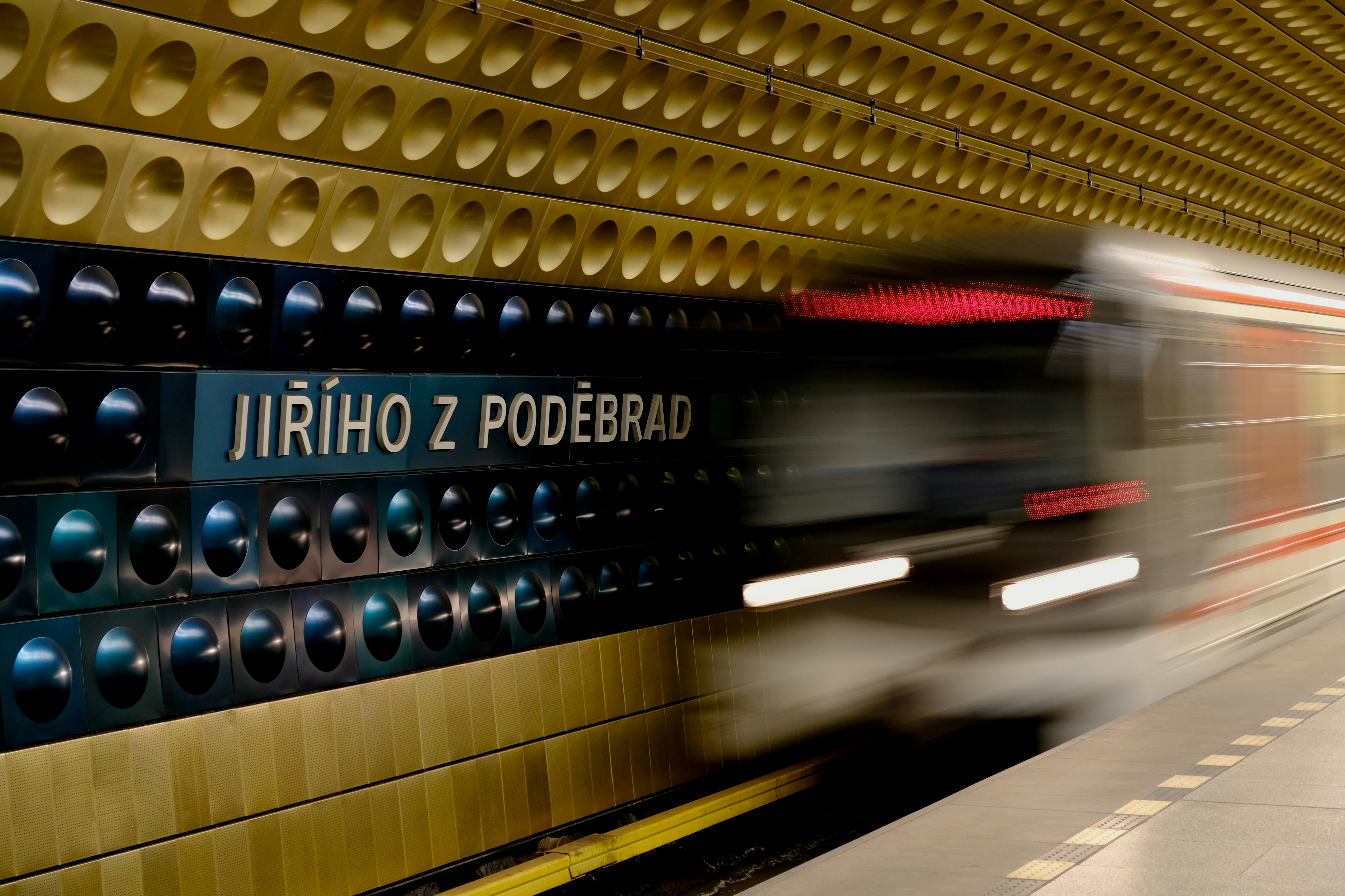 Jiřího z Poděbrad in Motion: Train Blur Across a Golden Platform Wall