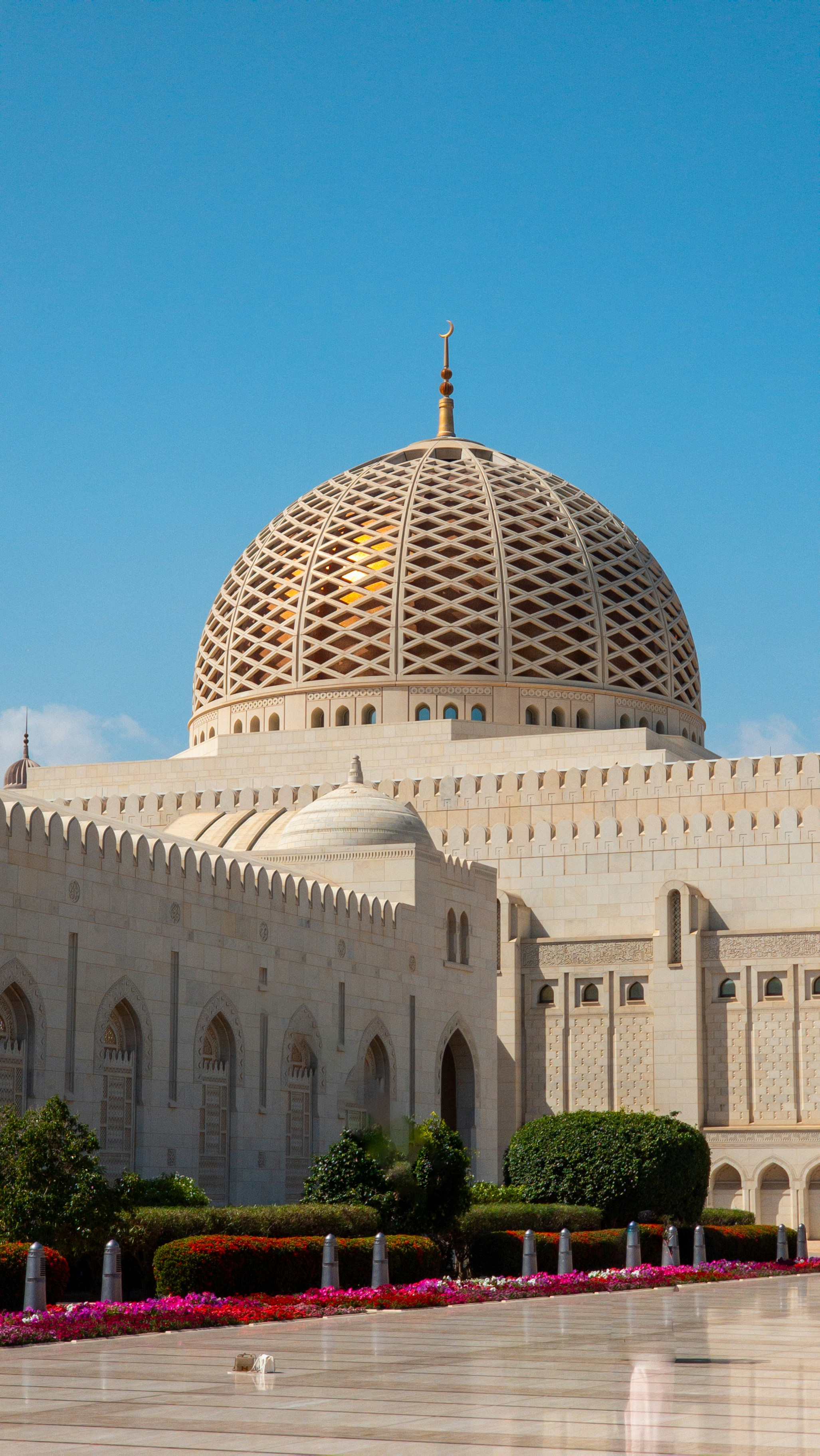 Intricate dome of a grand mosque, showcasing detailed geometric patterns against a clear blue sky. Lush gardens and vibrant flowers enhance the architectural beauty.