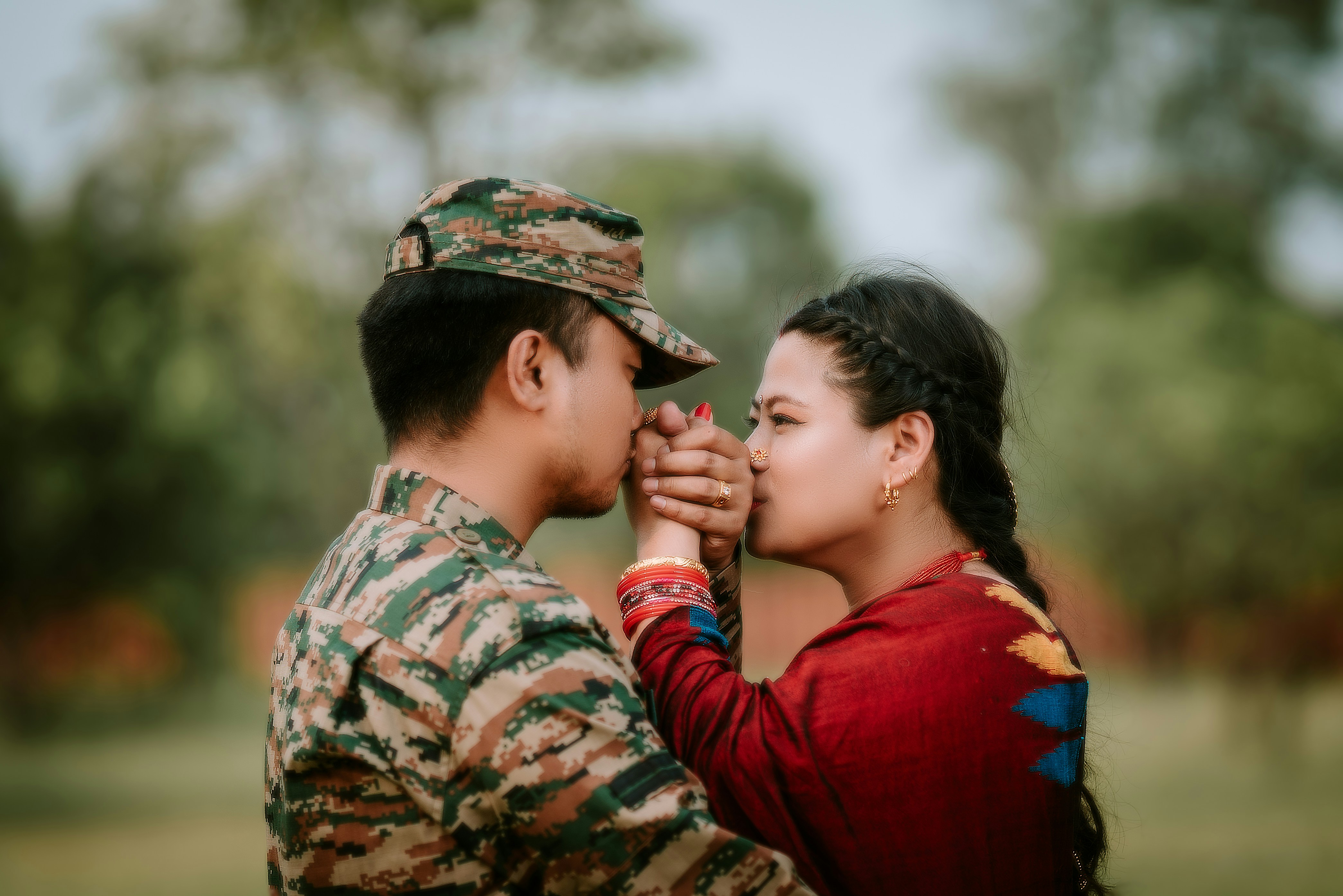 A soldier and a woman share an intimate moment, their foreheads touching as they hold hands, set against a blurred natural backdrop.