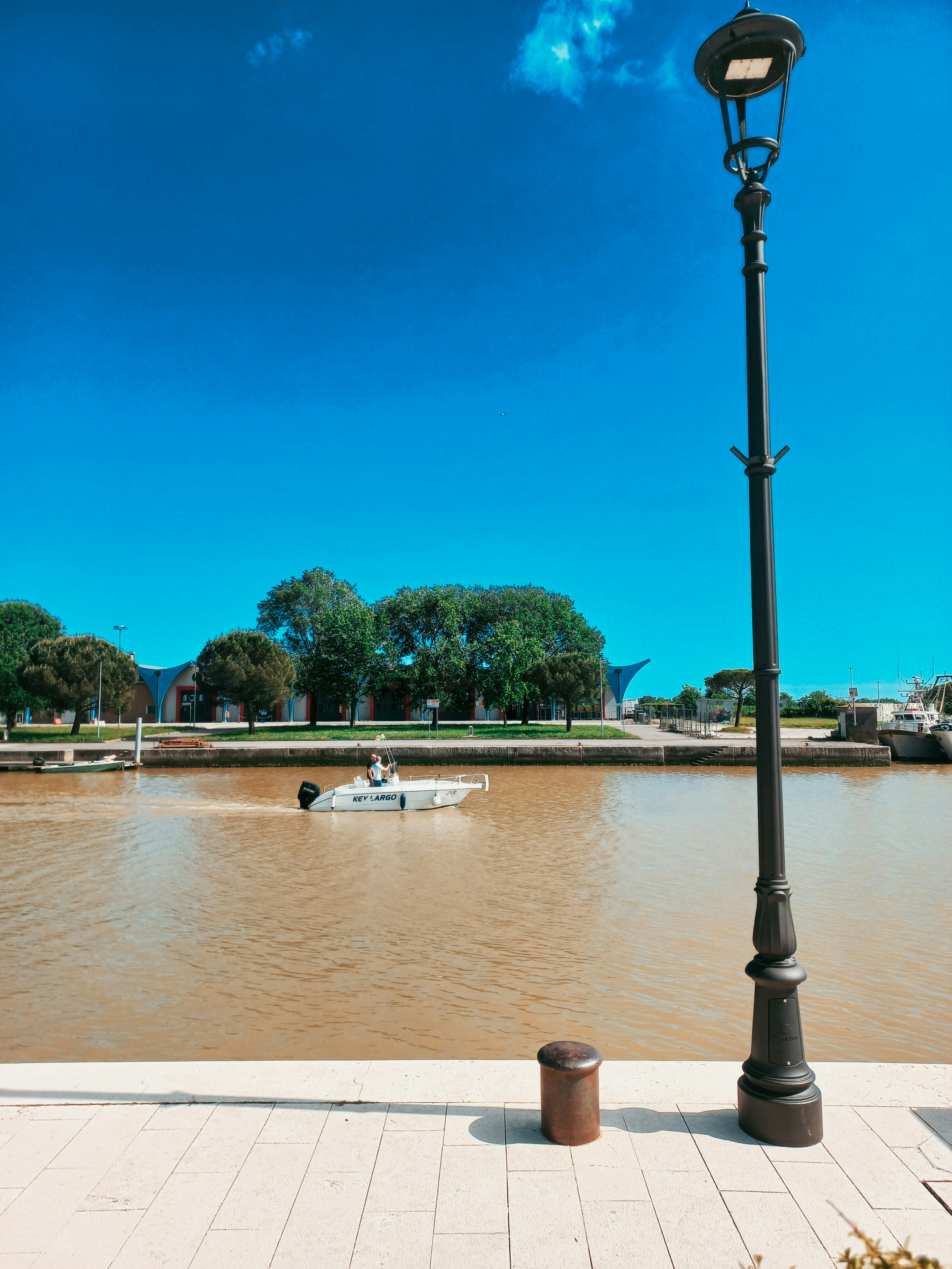 Riverside photograph showing a tall lamppost on the right, a small boat on brown water, and a row of trees beneath a bright blue sky.
