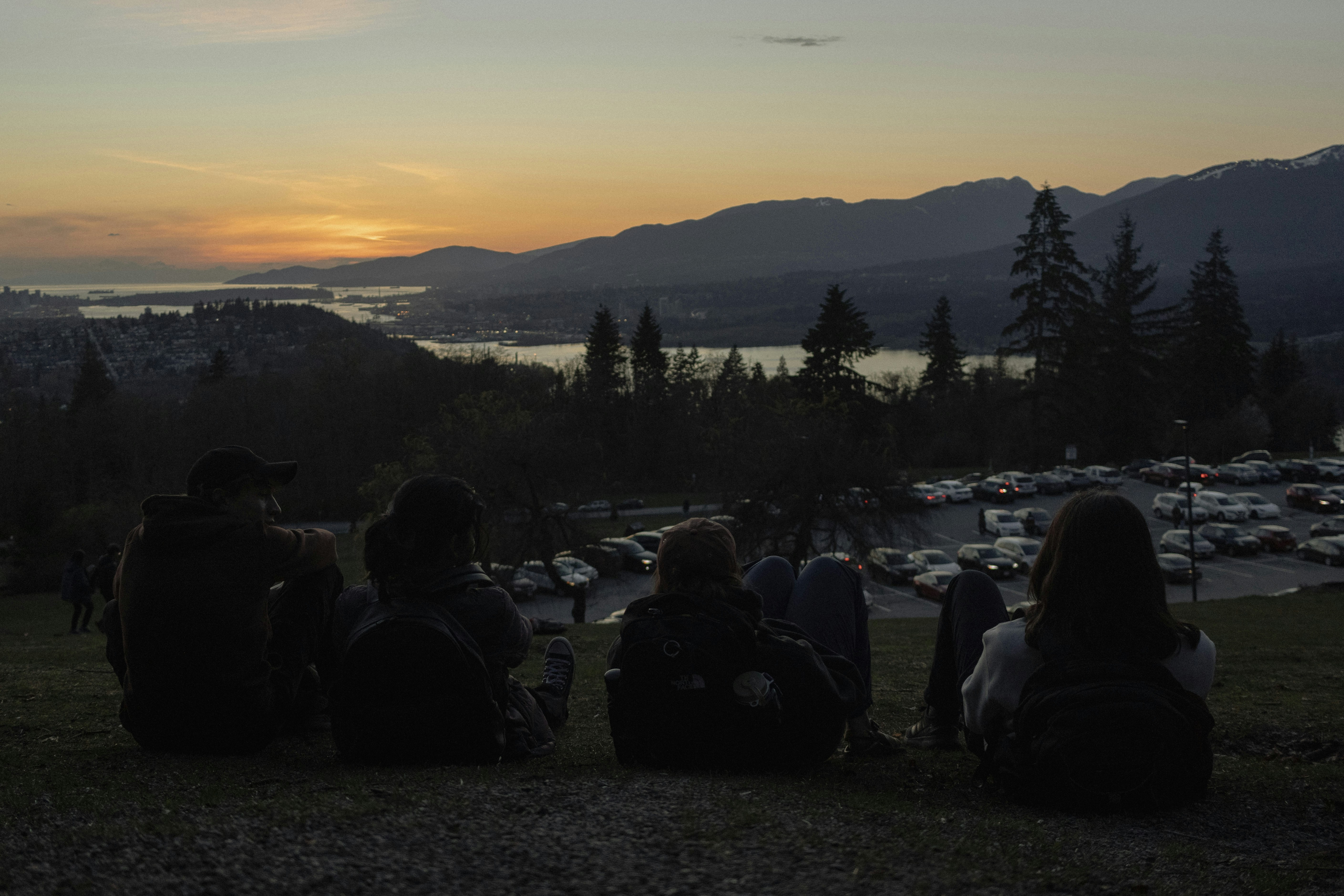 Silhouetted friends sitting on a hillside watching a sunset over distant mountains and a shimmering lake.