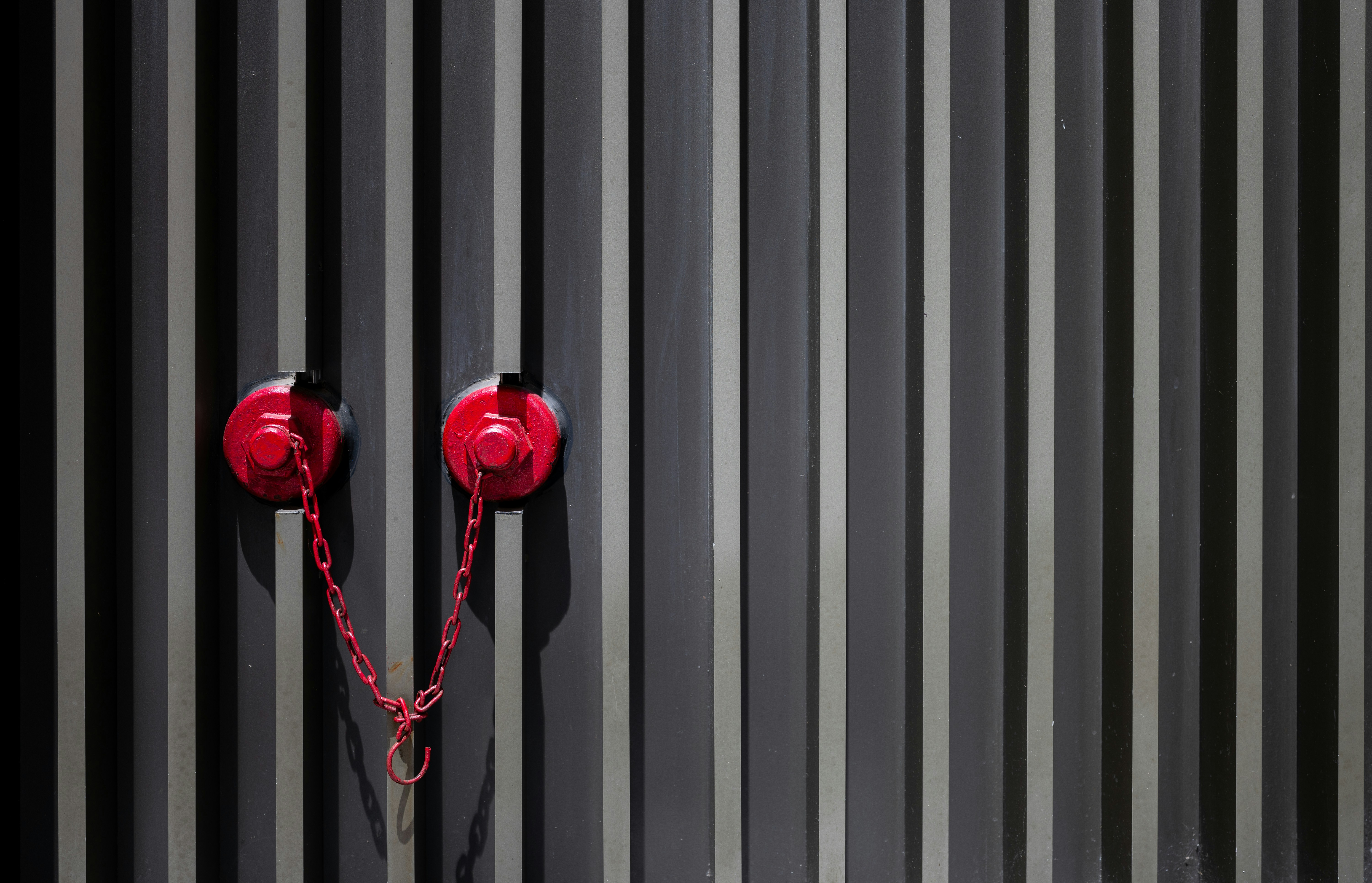 Two red circular latches connected by a chain are mounted on a vertical grey corrugated metal wall.