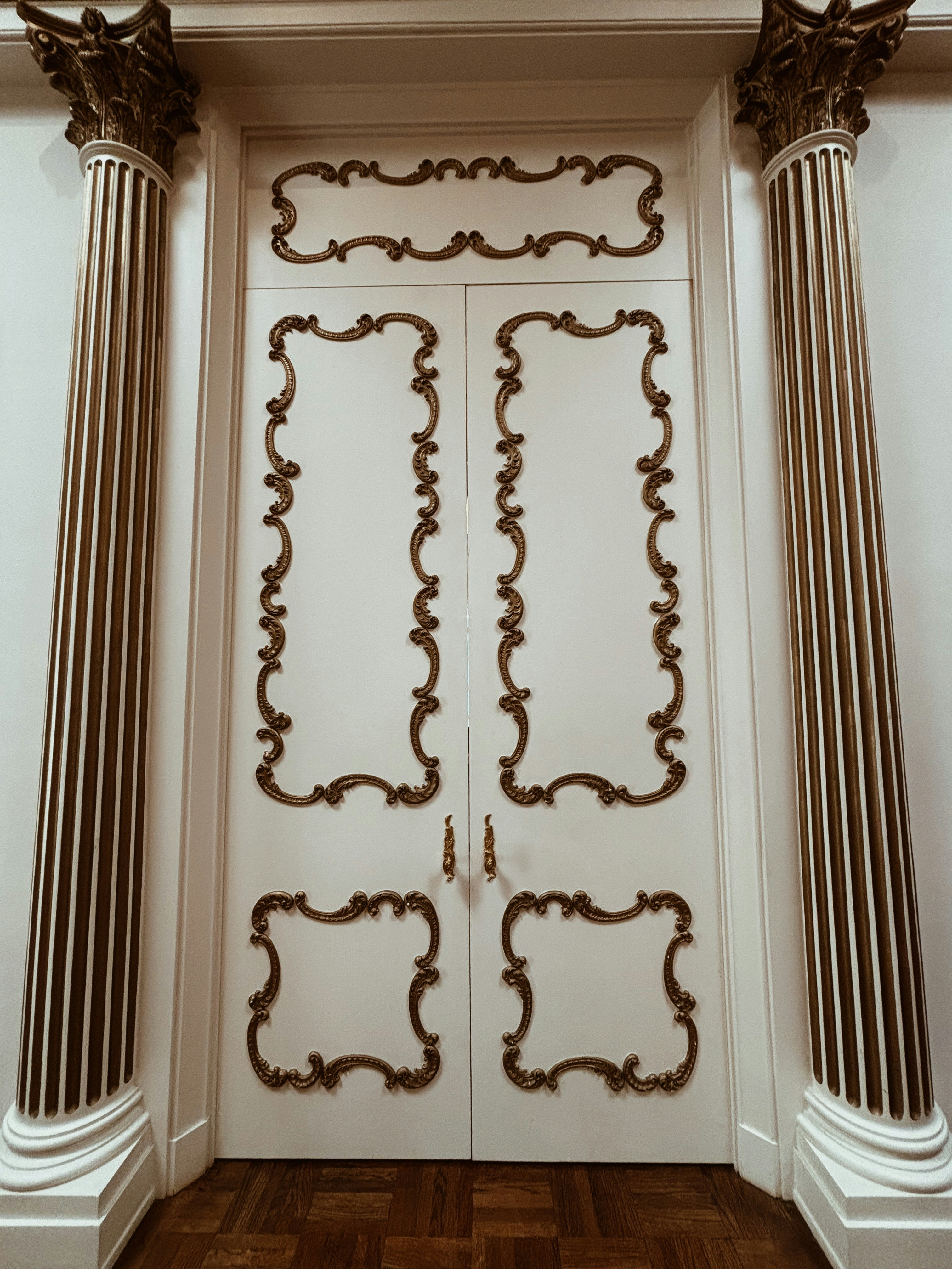 Double doors adorned with gold scrollwork framed by fluted columns in a neoclassical interior.
