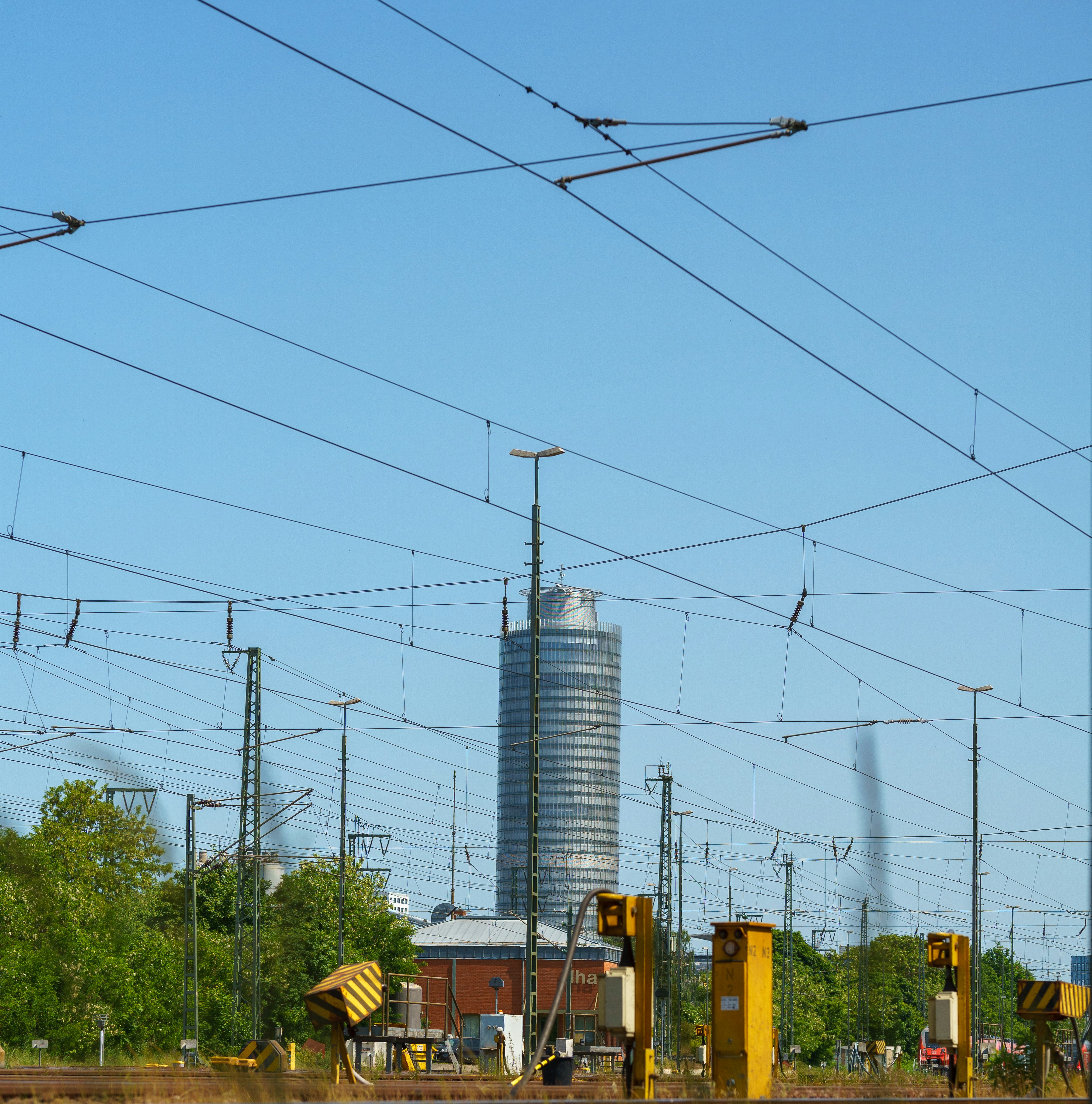 This image showcases an urban landscape dominated by a intricate network of power lines and utility poles against a clear blue sky. The prominent feature is a modern high-rise building with a distinctive glassy facade in the background, which appears to be a landmark structure in the area. In the foreground, there are several train cars or locomotives, painted in yellow and brown colors, suggesting an industrial or railway setting. The scene is framed by green foliage, likely trees, adding a natural element to the urban environment. The image captures the juxtaposition of modern architecture