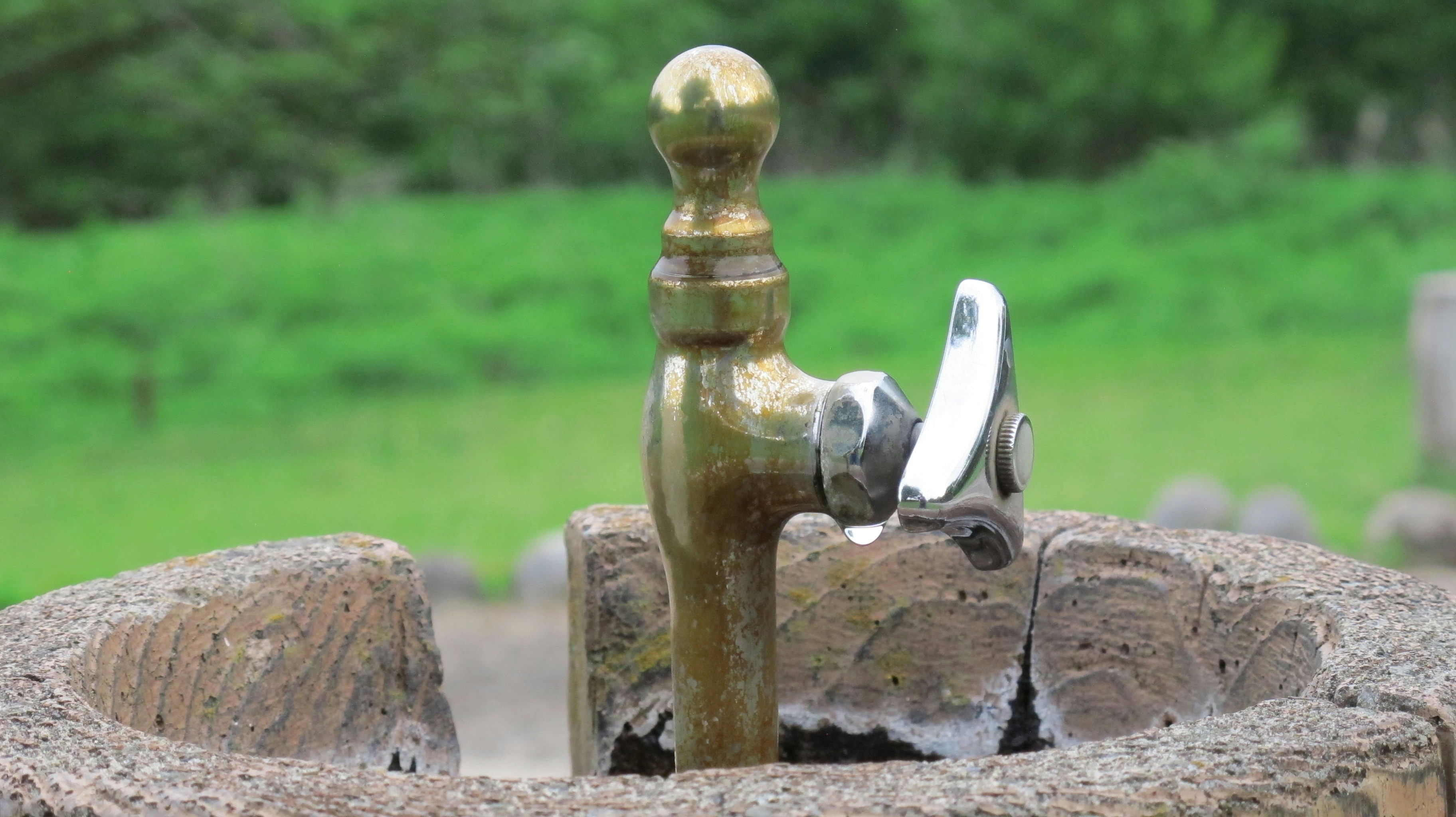 Close-up of a weathered brass spigot rising from a circular stone well, with a soft green garden backdrop.