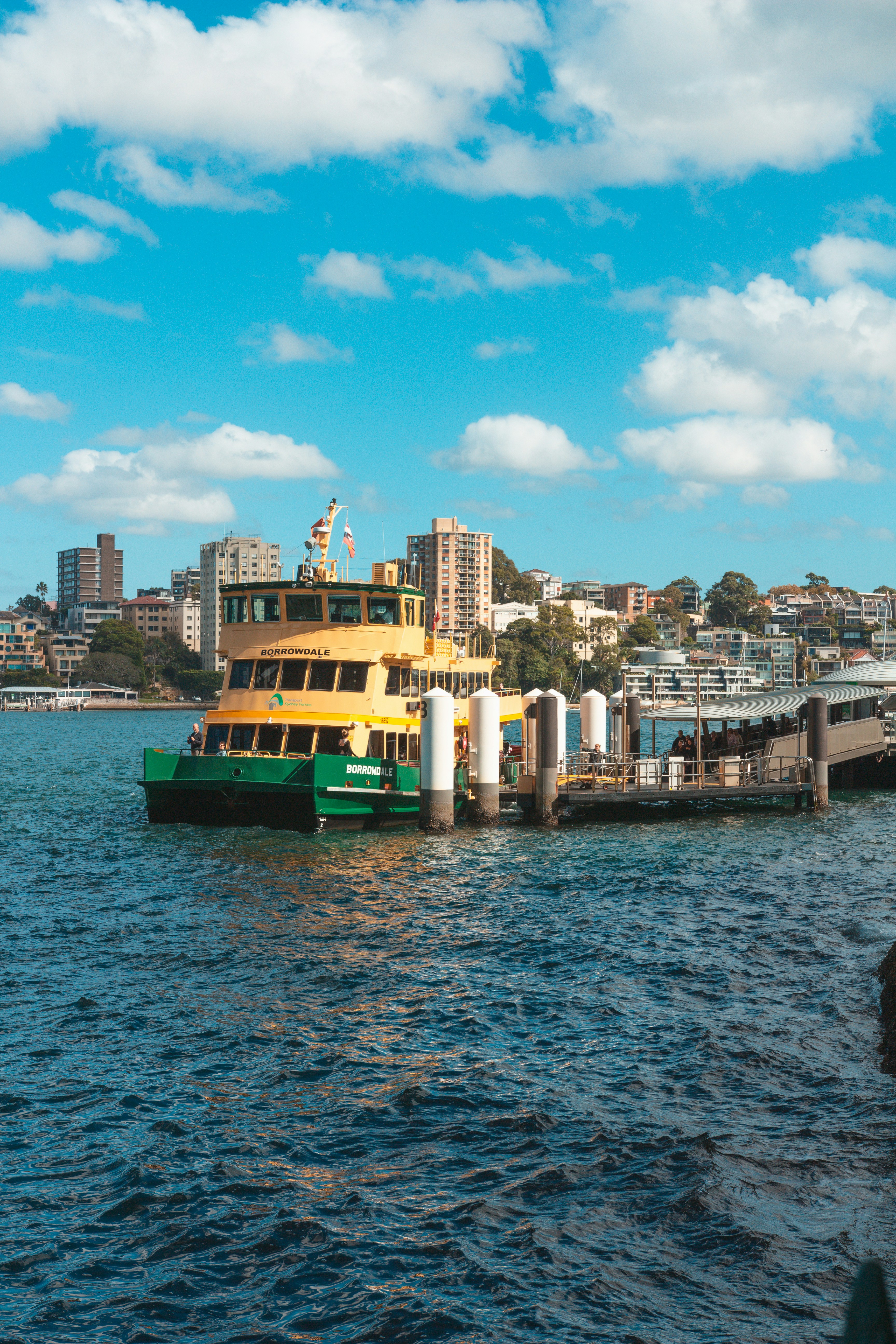 Green-Hulled Ferry Anchored at a Sunlit Harbor with Urban Backdrop
