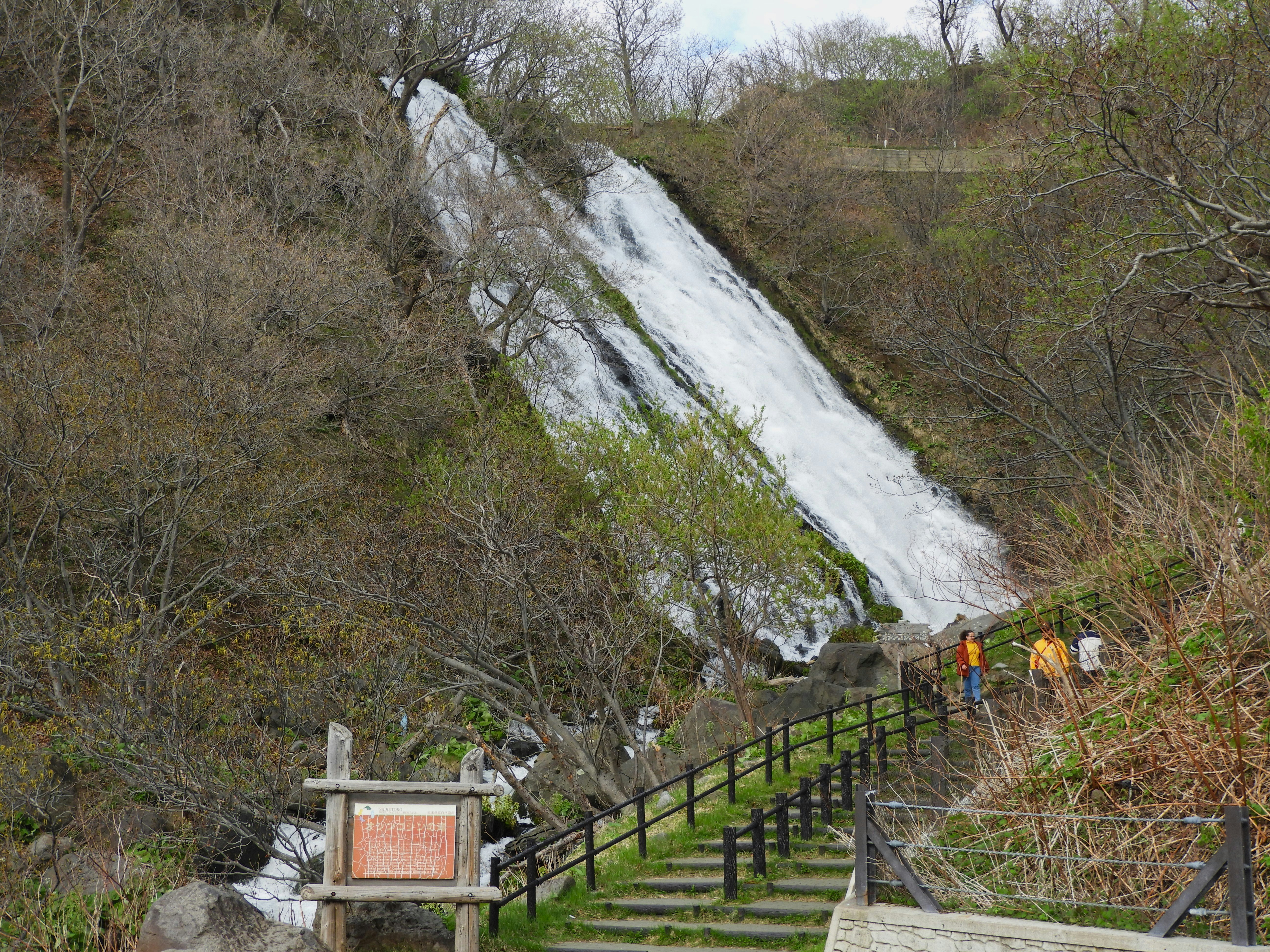 Photograph of a hillside waterfall cascading down a rocky slope, with a wooden railing trail. A few hikers observe the scene along the path.