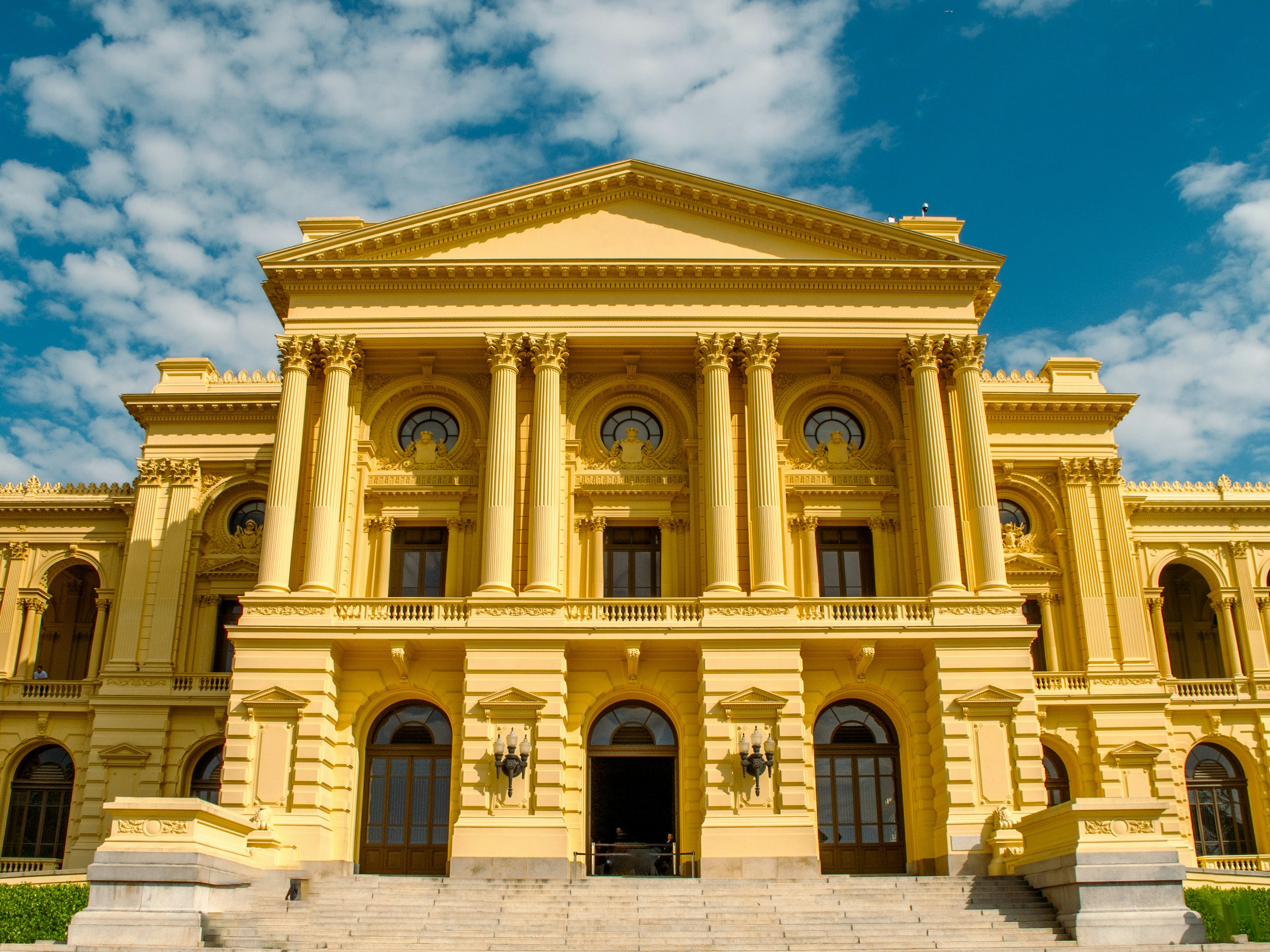 Large yellow neoclassical building with prominent columns and steps under a blue sky.
